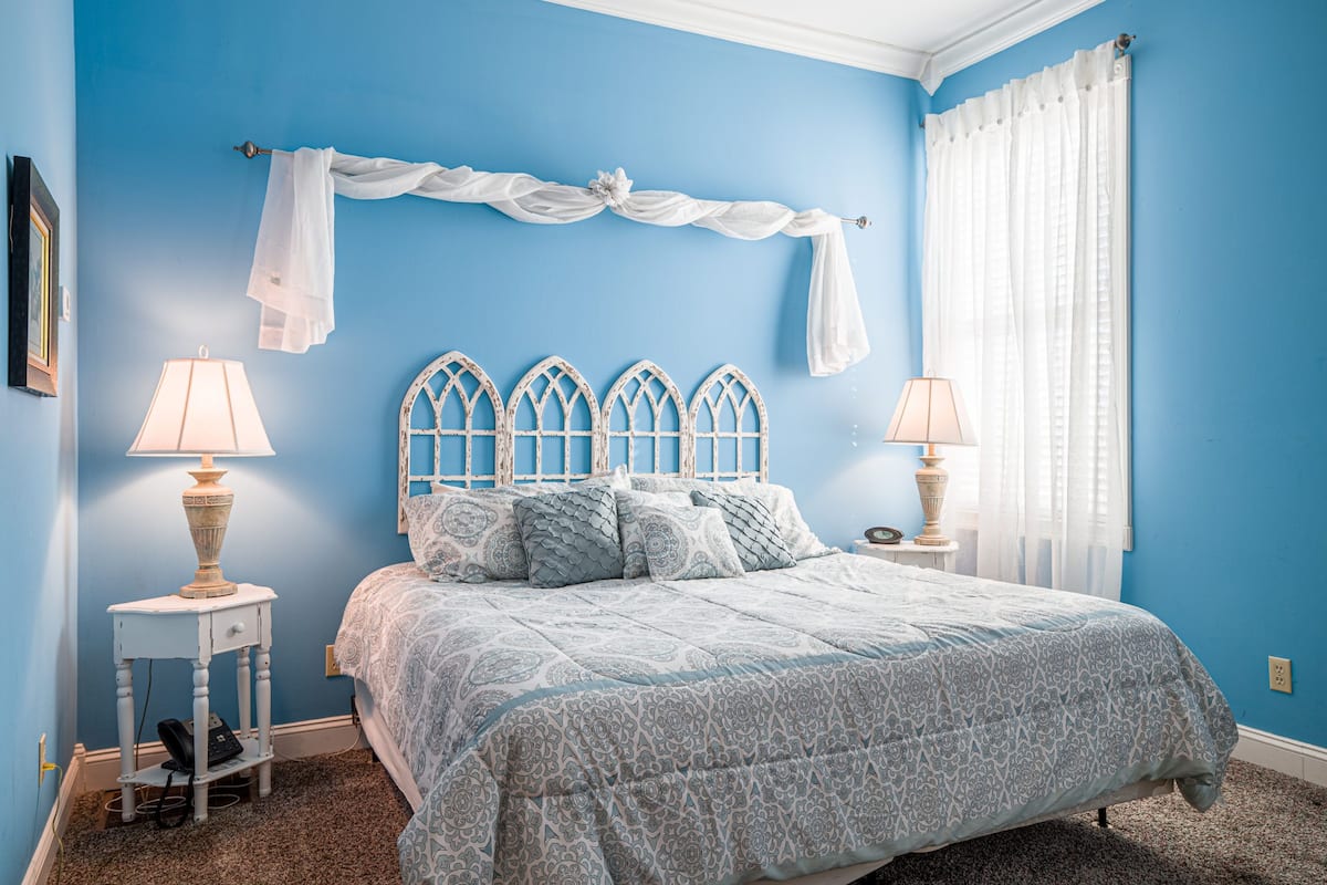 Quarto com parede azul, cama com colcha estampada, duas luminárias de cabeceira e cabeceira decorativa.