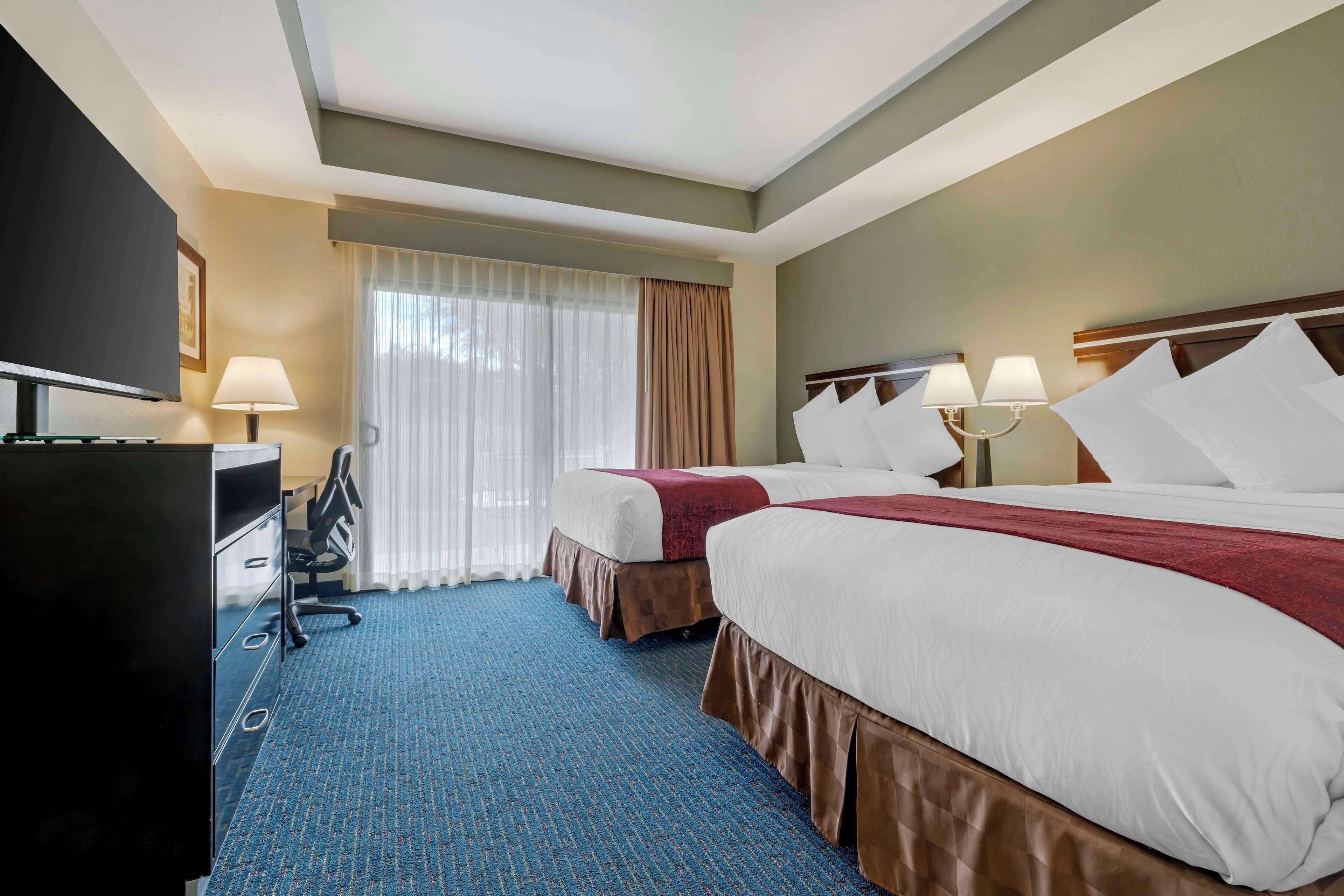 Quarto de hotel com duas camas, uma secretária e uma cadeira, televisão e janela com cortinas.