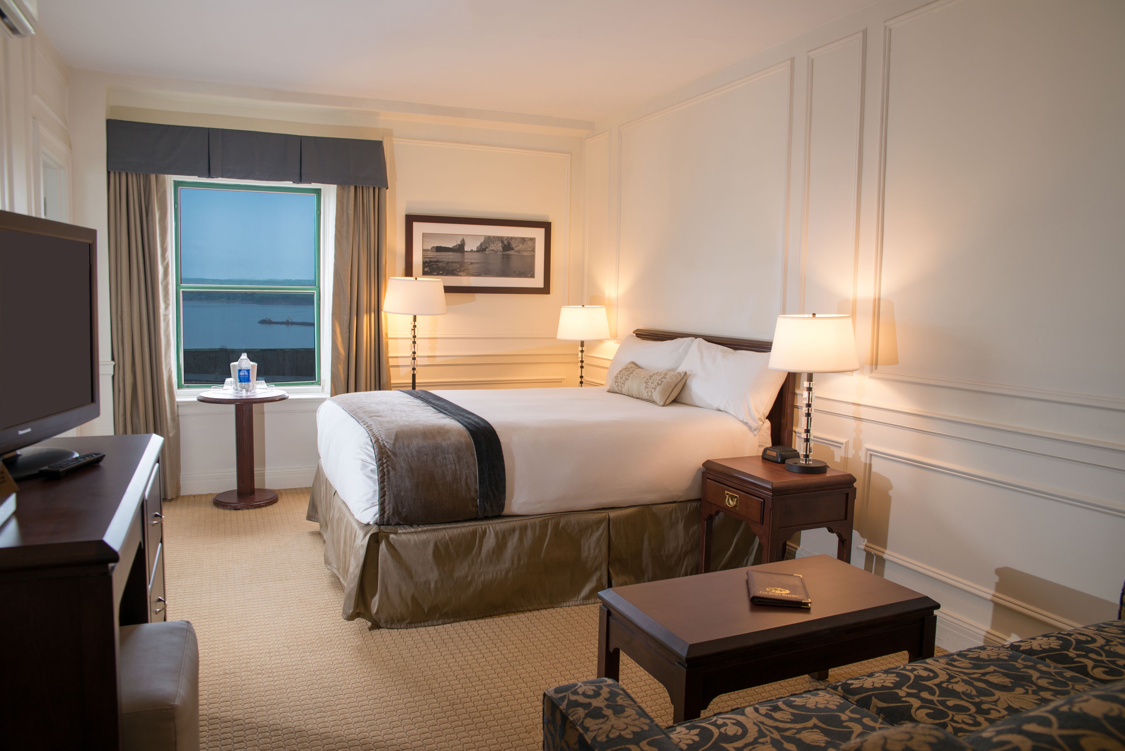 Quarto de hotel com cama, abajures, escrivaninha com televisão, mesinha e vista para o mar.