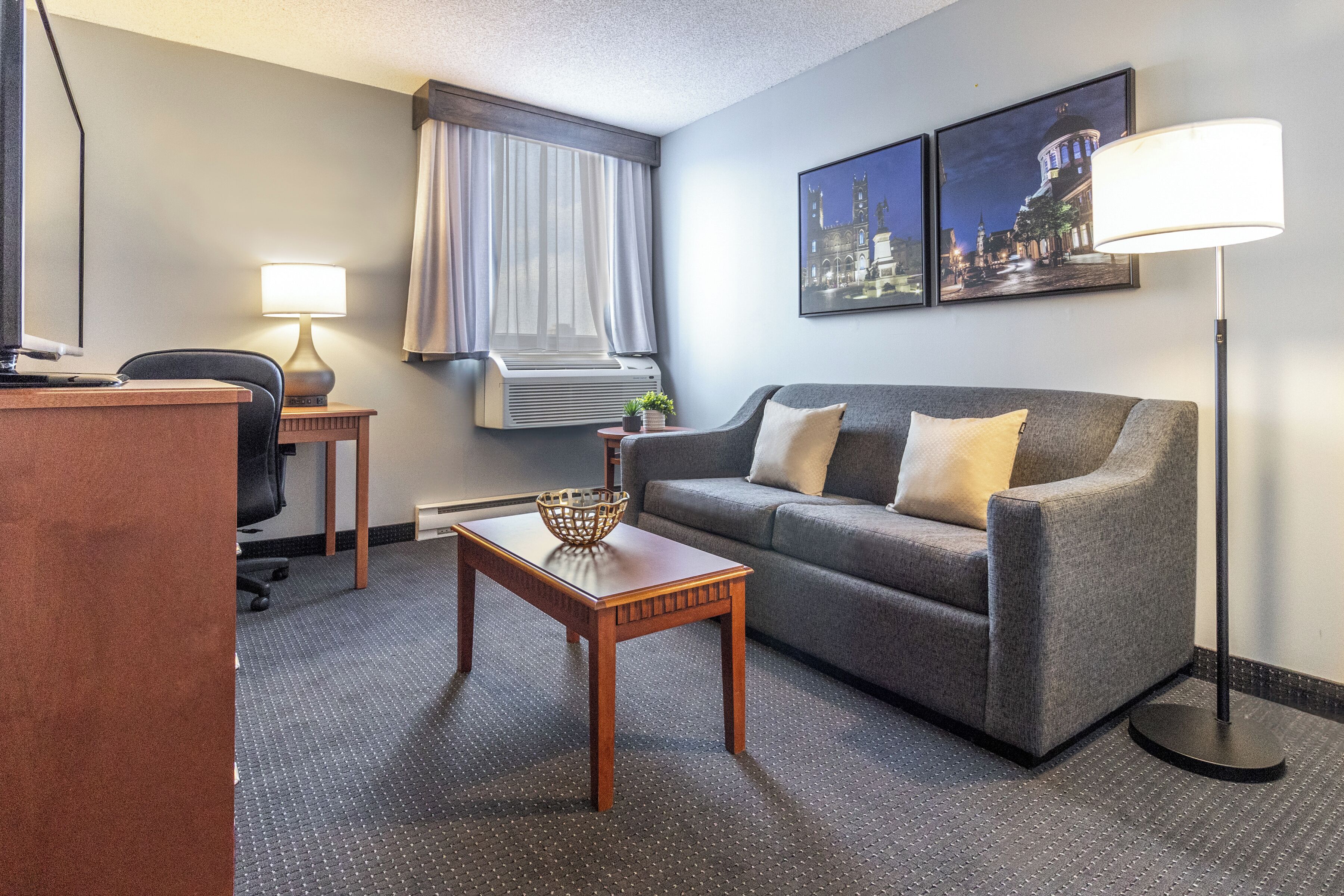 Quarto de hotel com um sofá cinzento, uma mesa de madeira com um cesto, uma escrivaninha com luminária e uma janela grande com cortinas.
