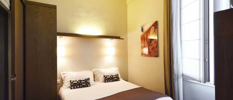 Quarto de hotel com cama, mesa de cabeceira, luminária, janela com cortinas e uma obra de arte pendurada na parede.