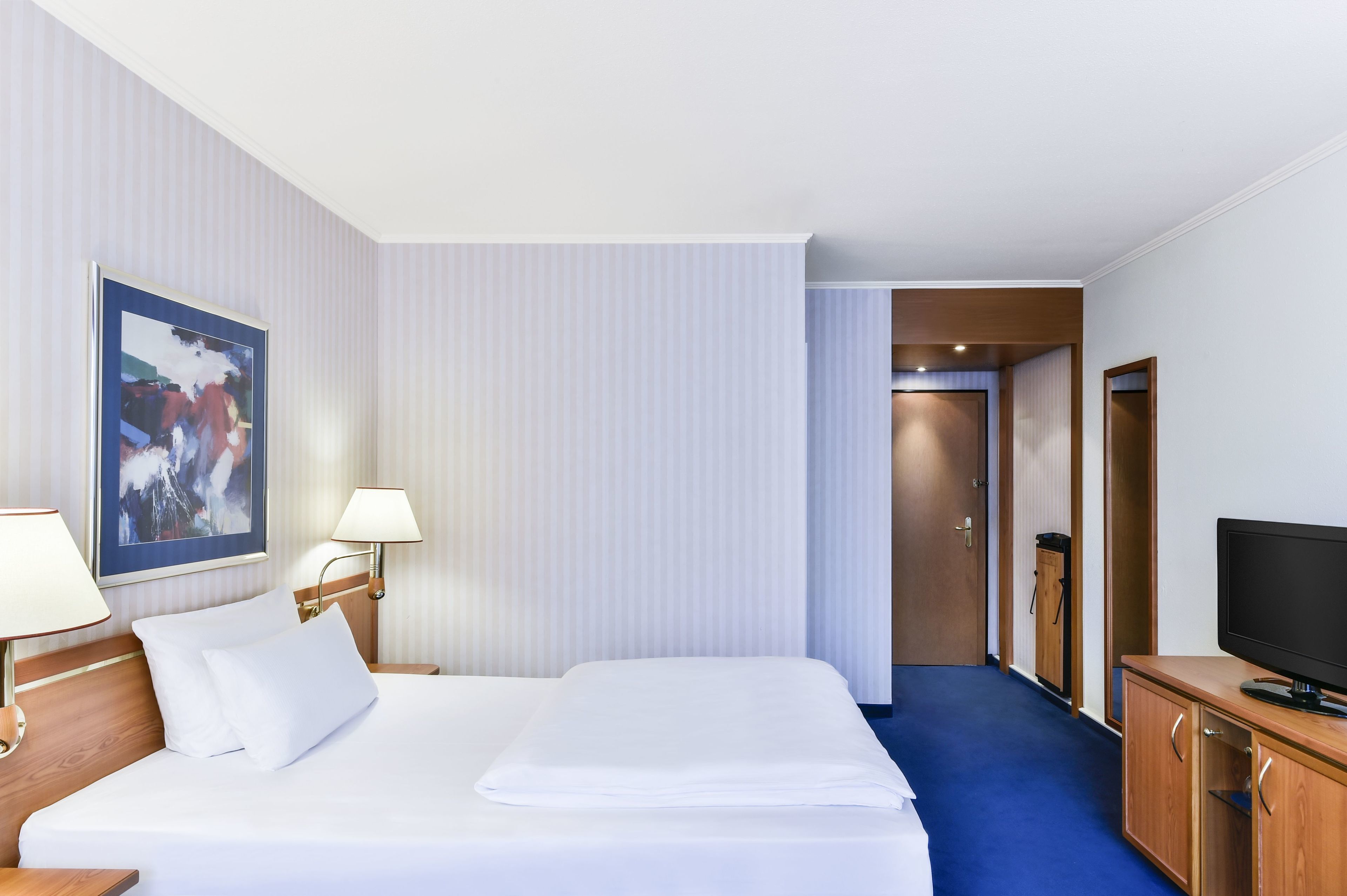 Quarto de hotel com uma cama grande, duas luminárias de cabeceira, uma televisão e um quadro pendurado na parede.