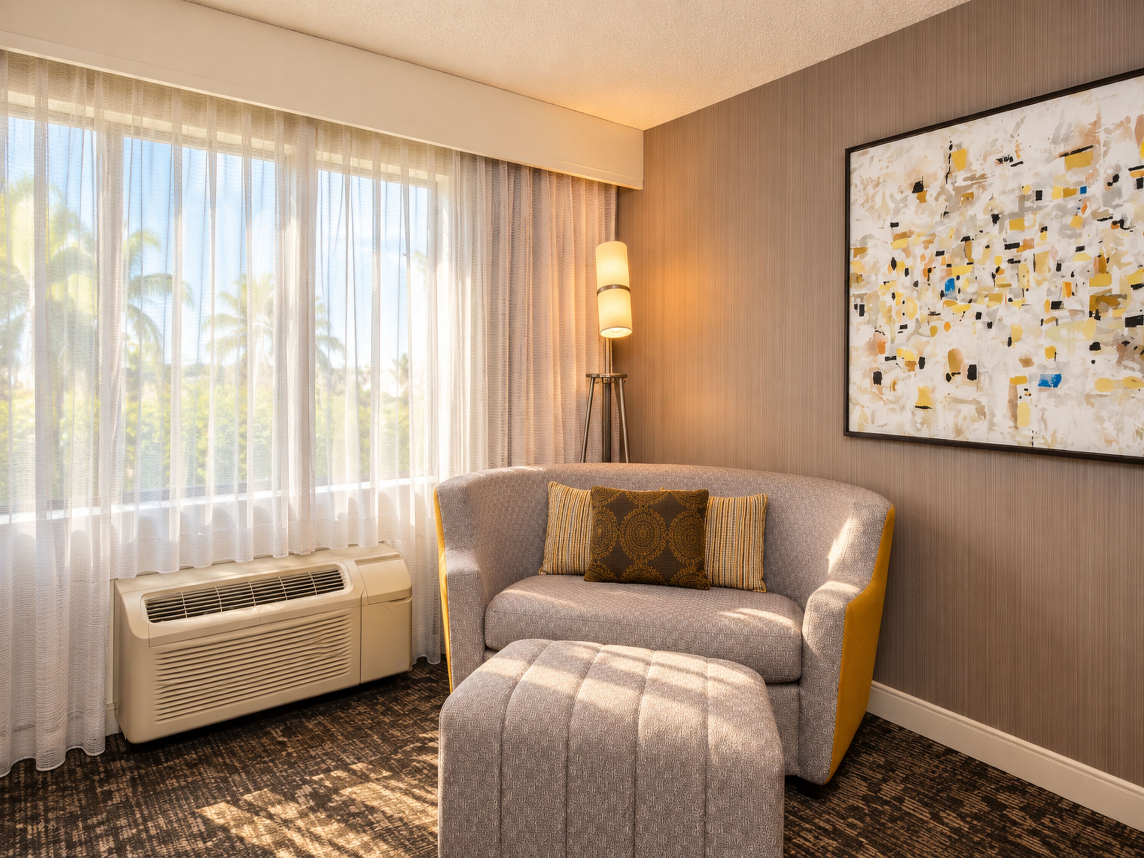 Quarto de hotel com uma janela grande, unidade de ar condicionado, sofá com almofada, luminária de chão e quadro abstrato na parede.