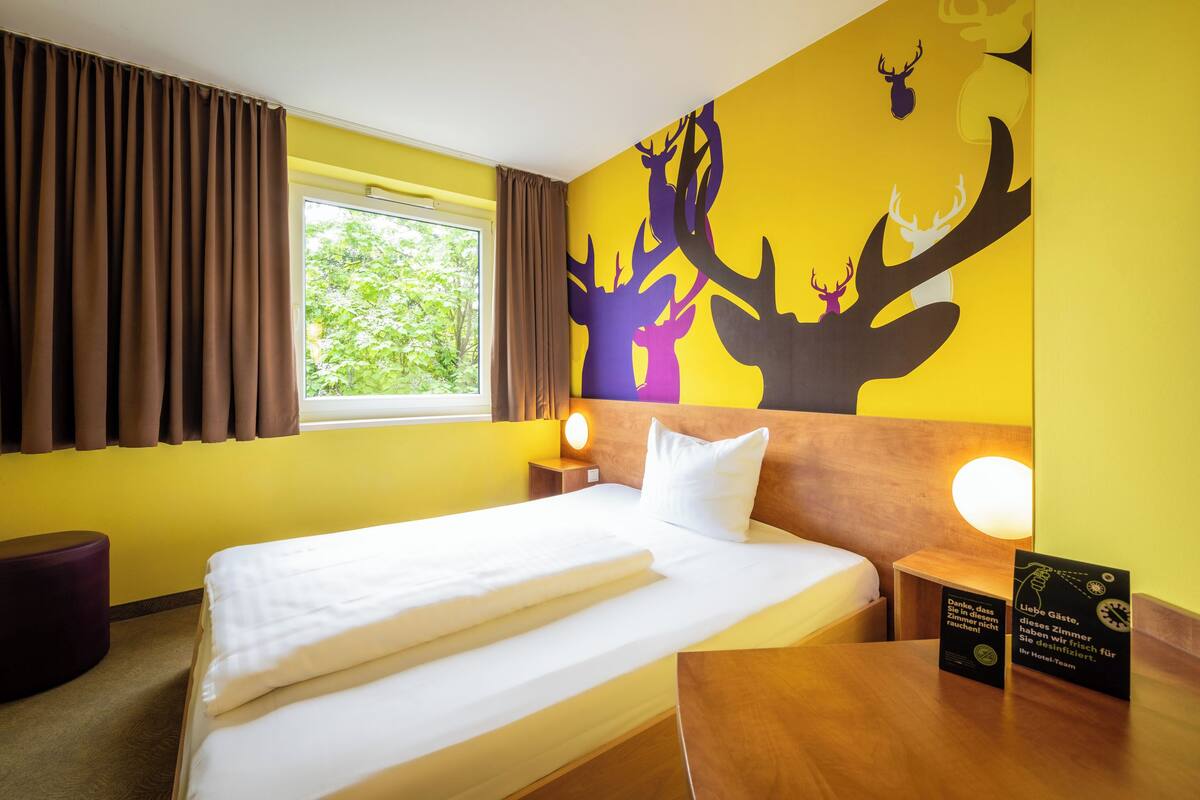 Quarto de hotel com cama, mesa de cabeceira, um mural na parede e janela com cortinas.