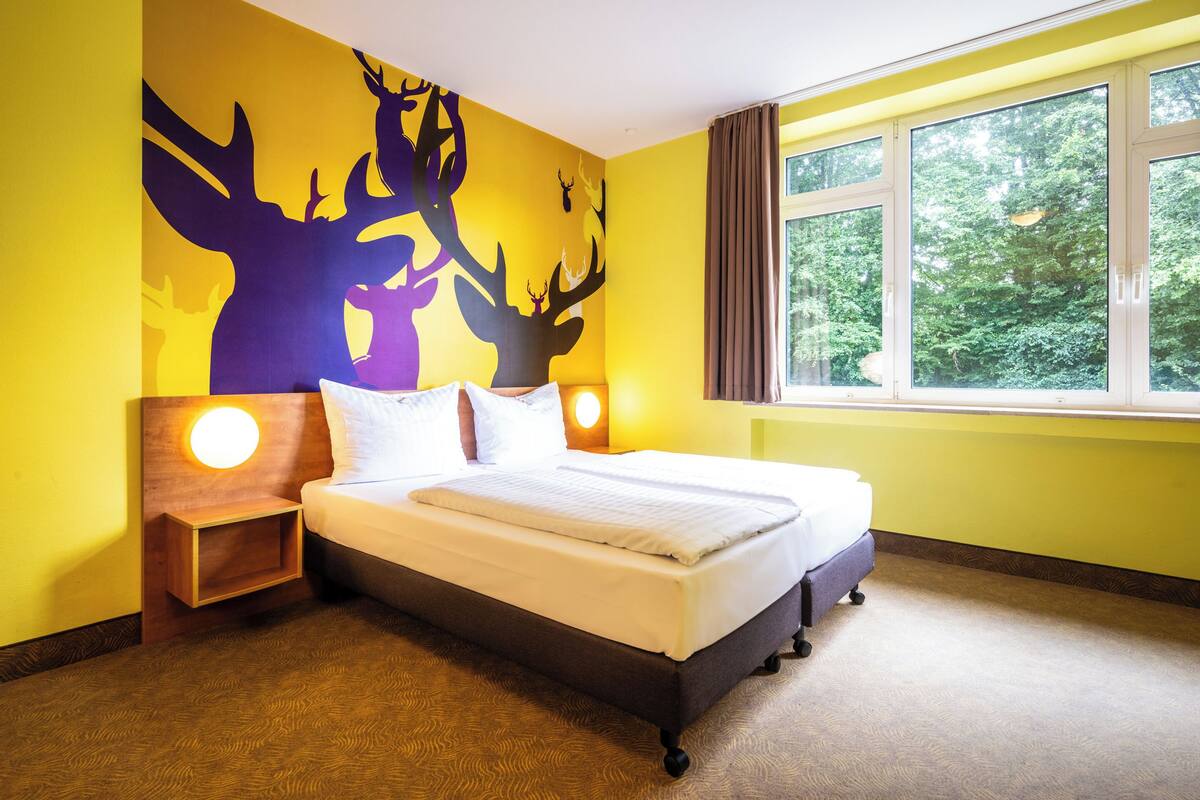 Quarto com cama, mesa de cabeceira, painel decorativo na parede e janela com cortinas.