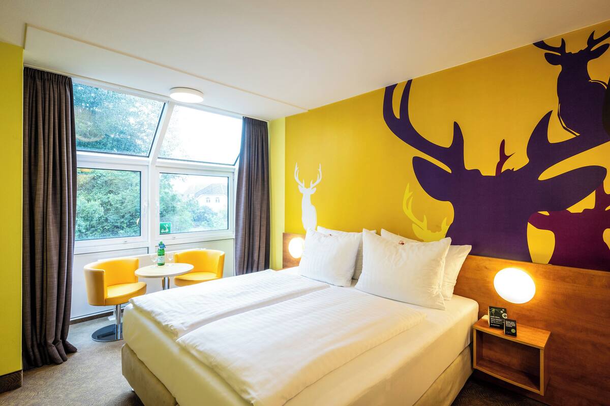 Quarto de hotel com uma cama grande, duas mesas de cabeceira, uma janela com cortinas, uma pequena mesa com cadeiras e um mural de veados na parede.