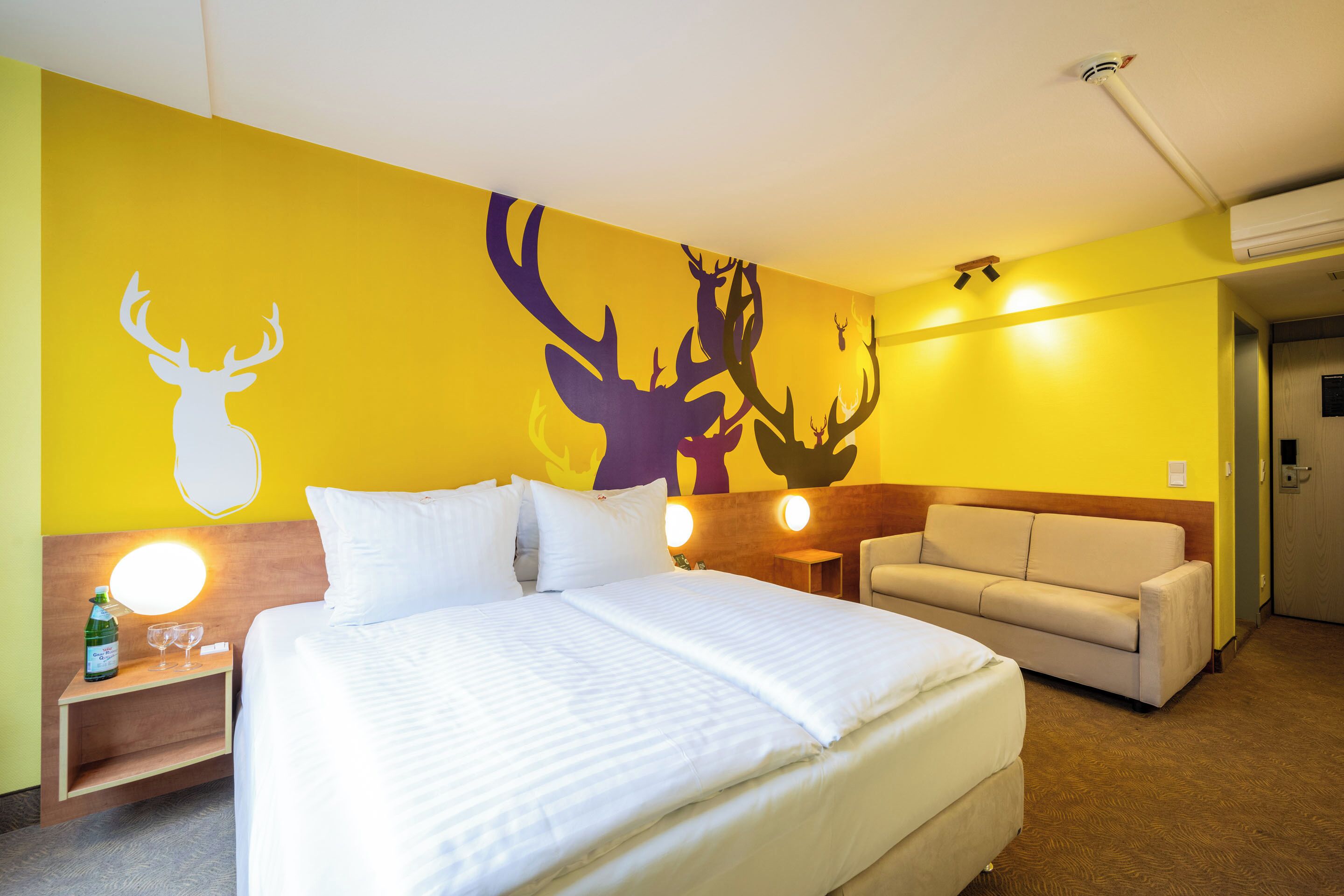 Quarto de hotel com uma cama grande, um sofá e um mural na parede com chifres de veado.