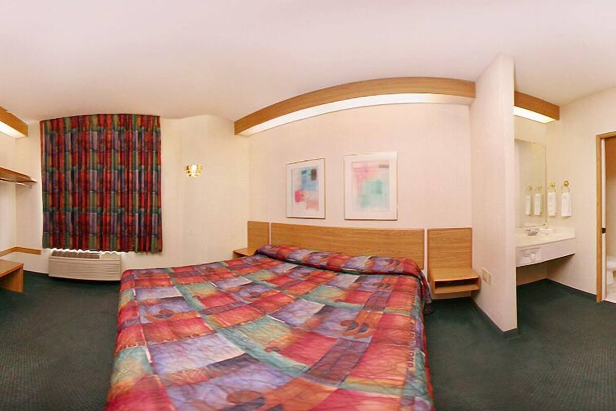 Quarto de hotel com uma cama grande, uma mesinha de cabeceira, uma cadeira, banheiro com pia e espelho, e janela com cortinas.