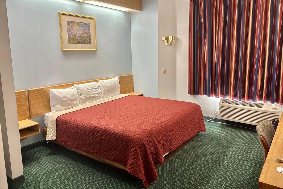 Quarto de hotel com cama, mesa de cabeceira, quadro na parede, janela com cortinas listradas e ar condicionado fixado na parede.