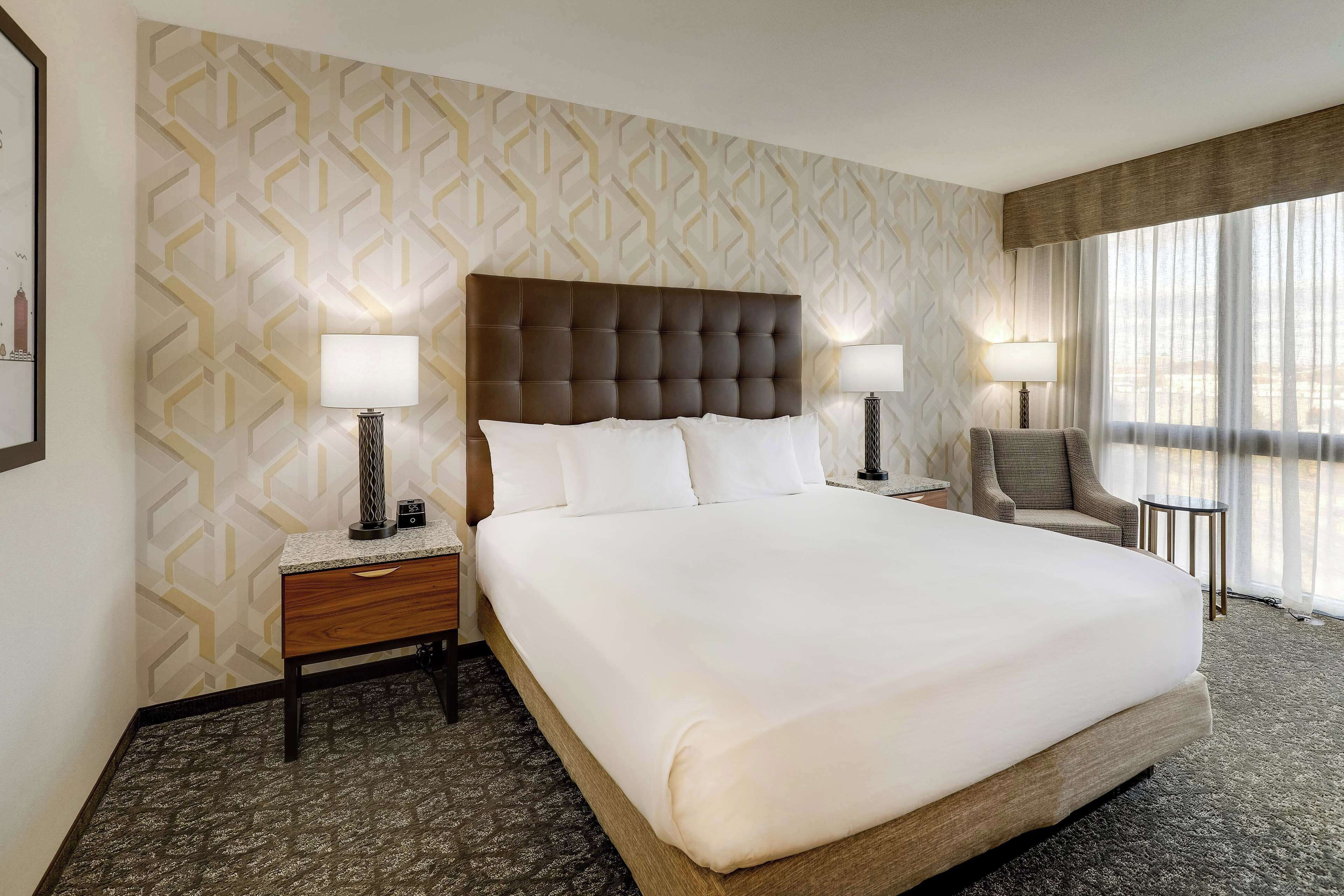 Quarto de hotel com uma cama grande, duas mesas de cabeceira com luminárias, uma cadeira e vista para o exterior através de amplas janelas.