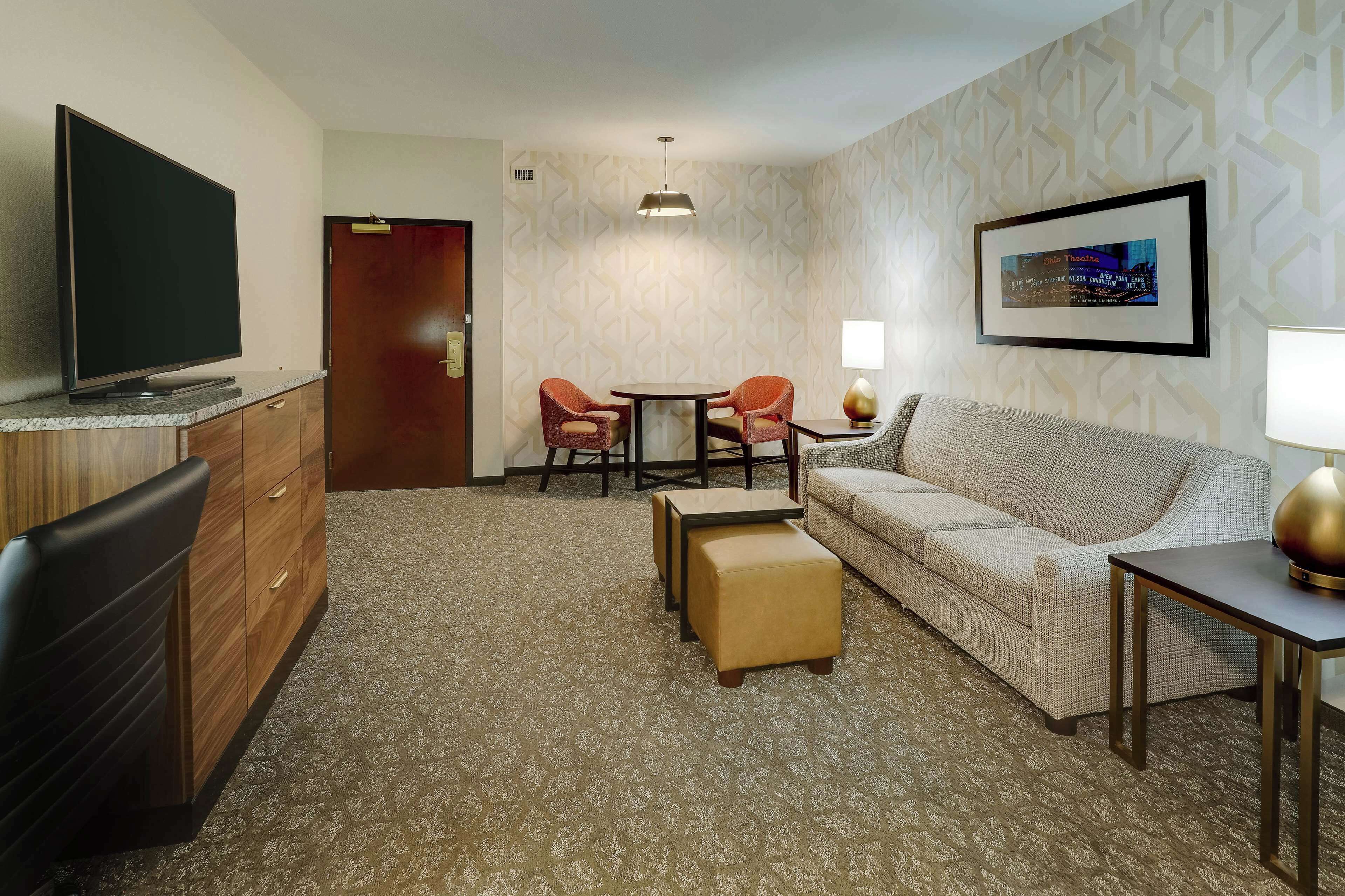 Quarto de hotel com televisão, sofá, mesa de jantar com cadeiras, um pequeno banquinho e um quadro na parede.