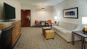Quarto de hotel com televisão, sofá, mesa de jantar com cadeiras, um pequeno banquinho e um quadro na parede.