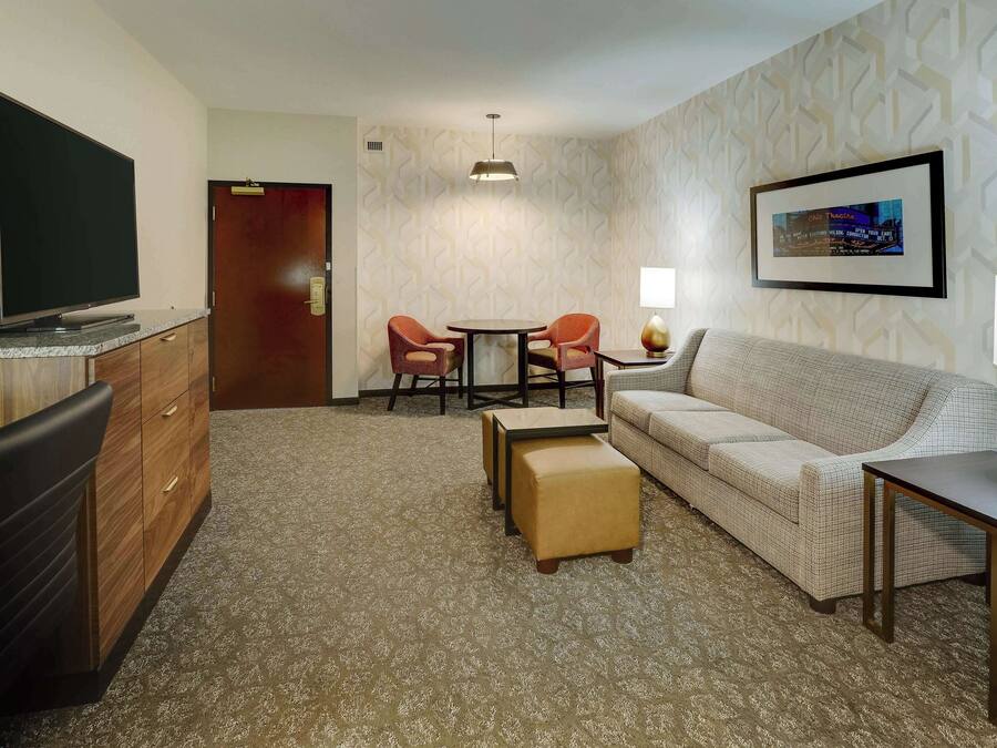 Quarto de hotel com televisão, sofá, mesa de jantar com cadeiras, um pequeno banquinho e um quadro na parede.
