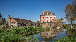 Um edifício histórico com fachada de pedra e persianas vermelhas, localizado ao lado de um corpo d'água calmo e vegetação exuberante.