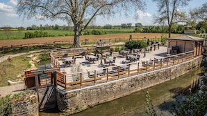 Um restaurante à beira-rio com área externa, uma ponte de pedra e vista para o rio e campos circundantes.