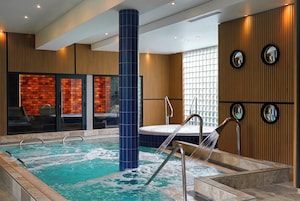 Uma piscina interior moderna com hidromassagem, painéis de madeira e uma parede de vidro com padrão de tijolos.