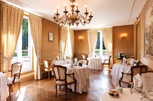 Uma sala de jantar com várias mesas preparadas para o serviço, elegantes lustres e cadeiras de madeira clássicas.