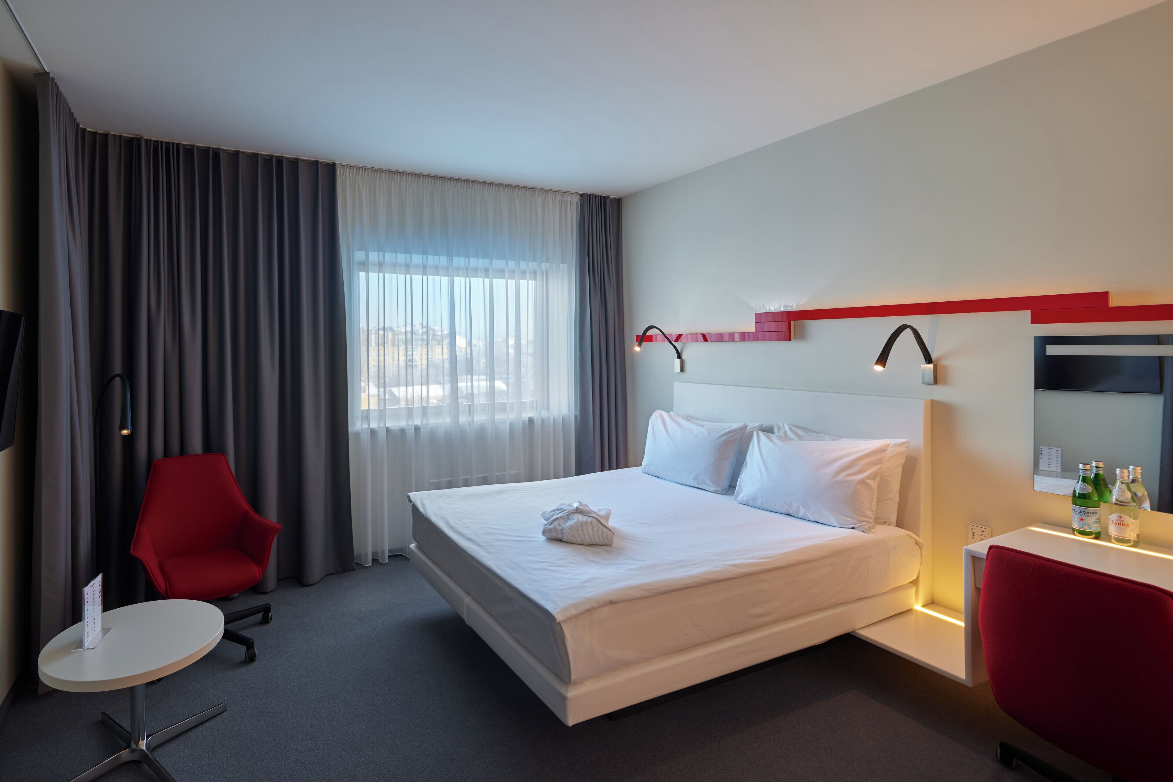 Quarto de hotel moderno com uma cama grande, uma cadeira vermelha, uma mesa pequena e uma escrivaninha com abajur.