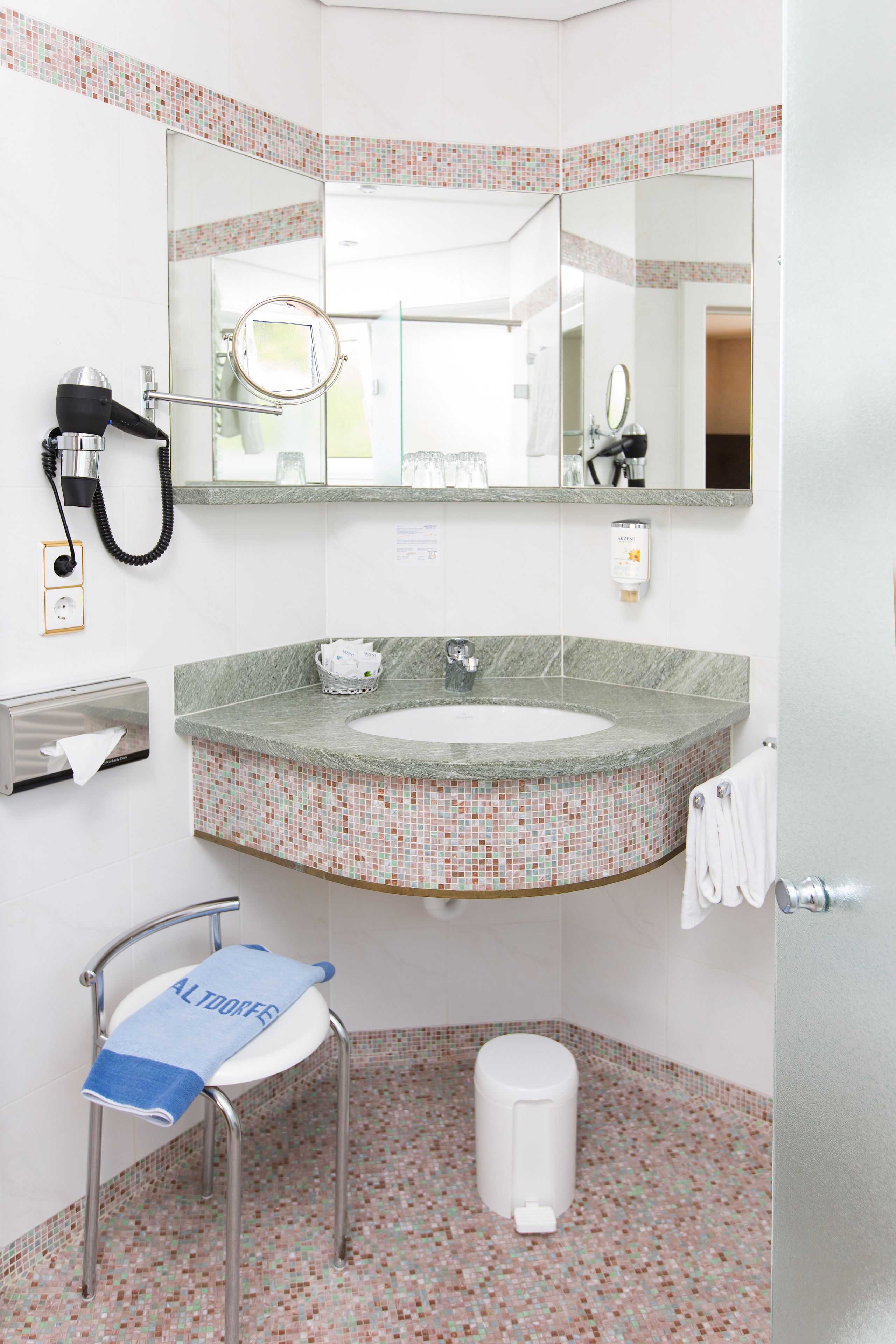 Um banheiro com piso em mosaico, pia com espelho acima, porta-toalhas e secador de cabelo em suporte fixado na parede.