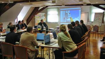 Sala de conferência com tela de projeção, vigas de madeira no teto e participantes sentados em mesas com laptops.