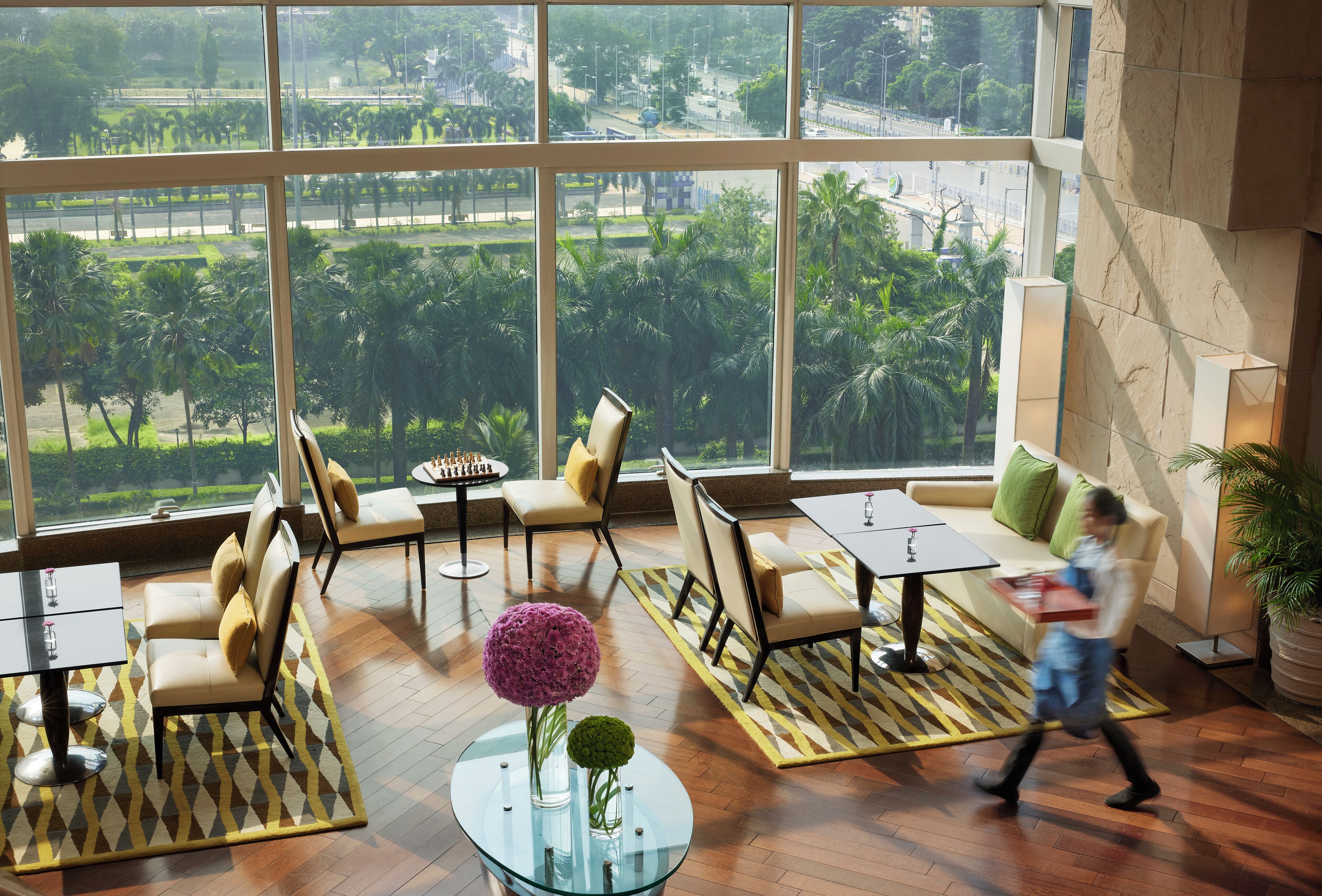 Um saguão moderno de hotel, com amplas janelas, assentos confortáveis e uma mesa de centro de vidro com flores decorativas.
