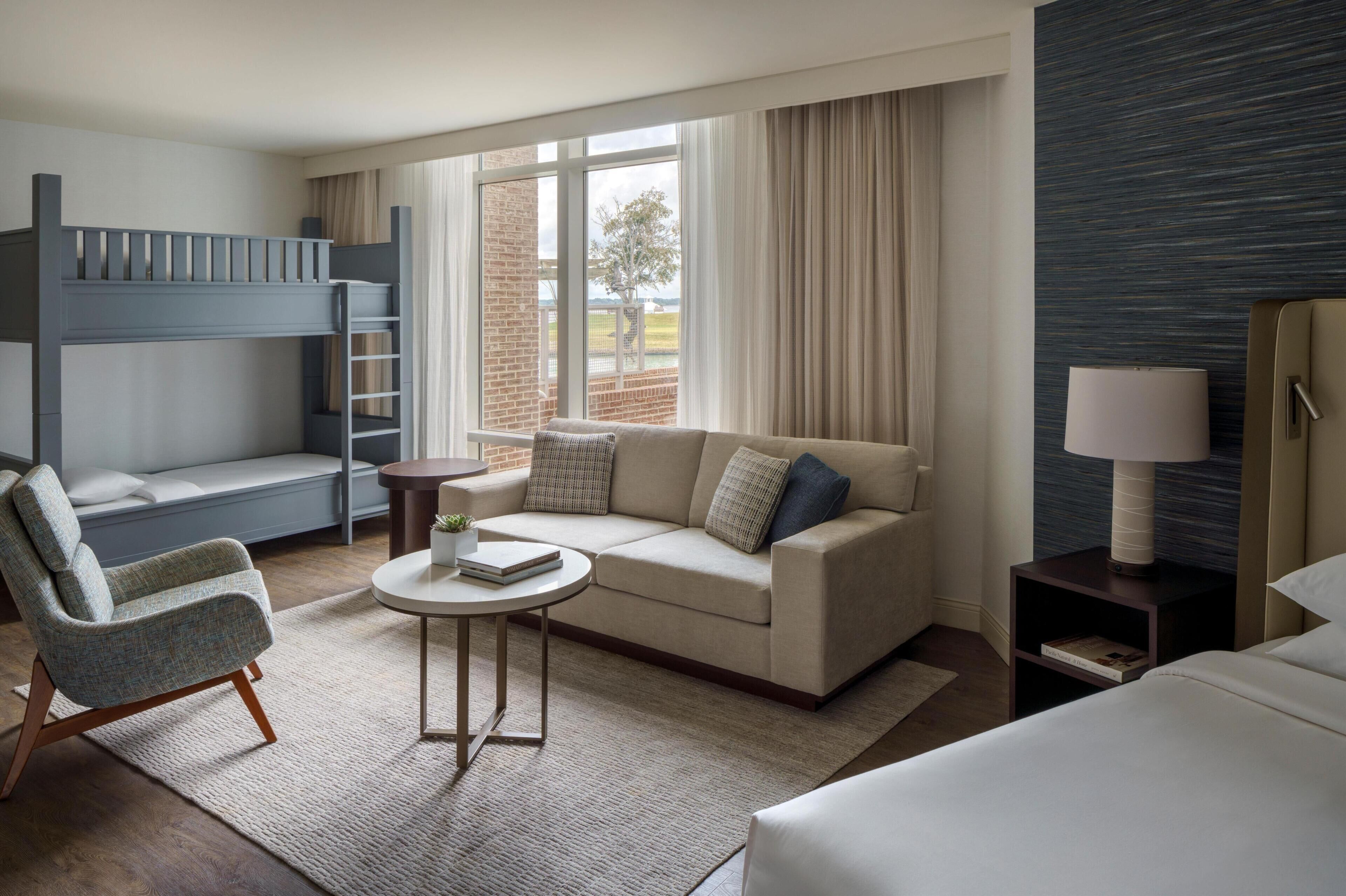 Quarto de hotel com uma cama beliche, um sofá, uma mesa de centro redonda, uma cadeira, uma luminária e vista para o exterior.