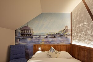 Quarto de hotel com um mural grande de uma ponte e edifícios, uma cabeceira de madeira, uma cadeira azul com bolinhas e uma cama com roupa de cama branca e um travesseiro azul.