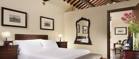Uma cama bem arrumada com cabeceira de madeira, mesa de cabeceira com abajur e um banco. Há quadros nas paredes e um espelho sobre a cama.