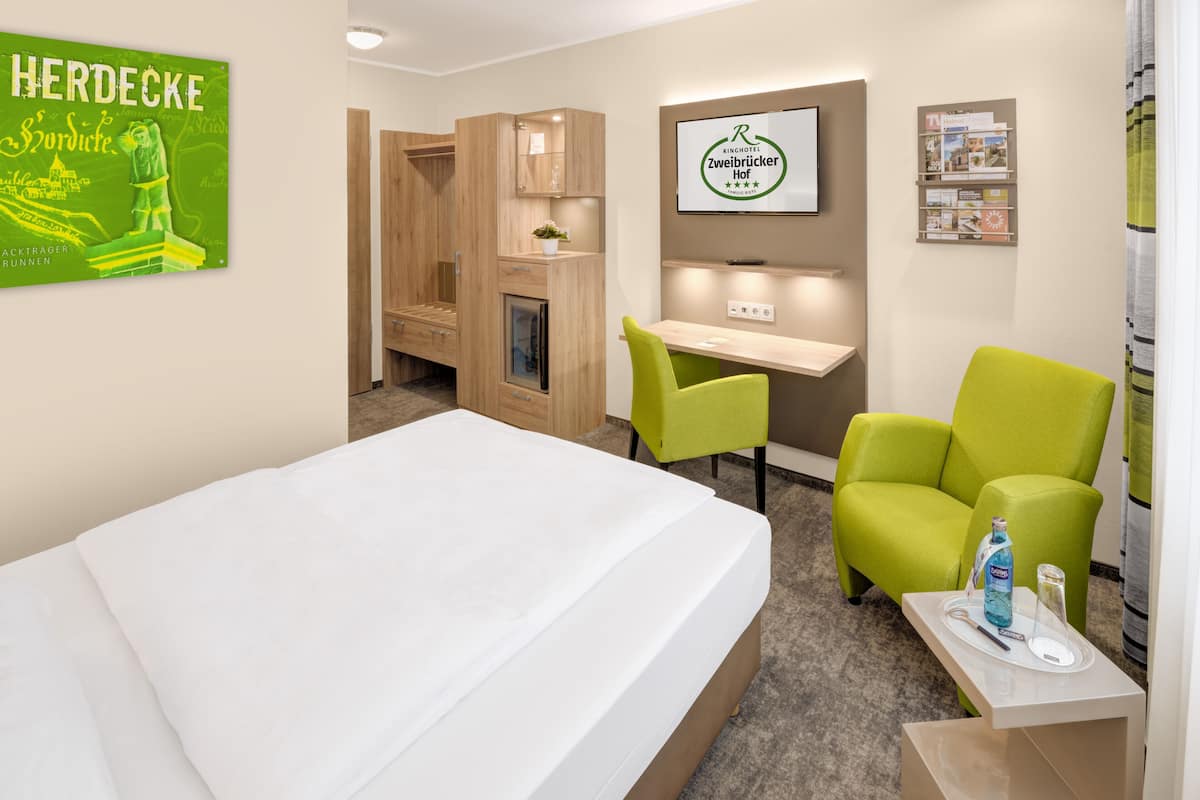 Quarto de hotel com cama, escrivaninha, cadeira, uma mesinha com garrafa e copo, TV na parede e um quadro.