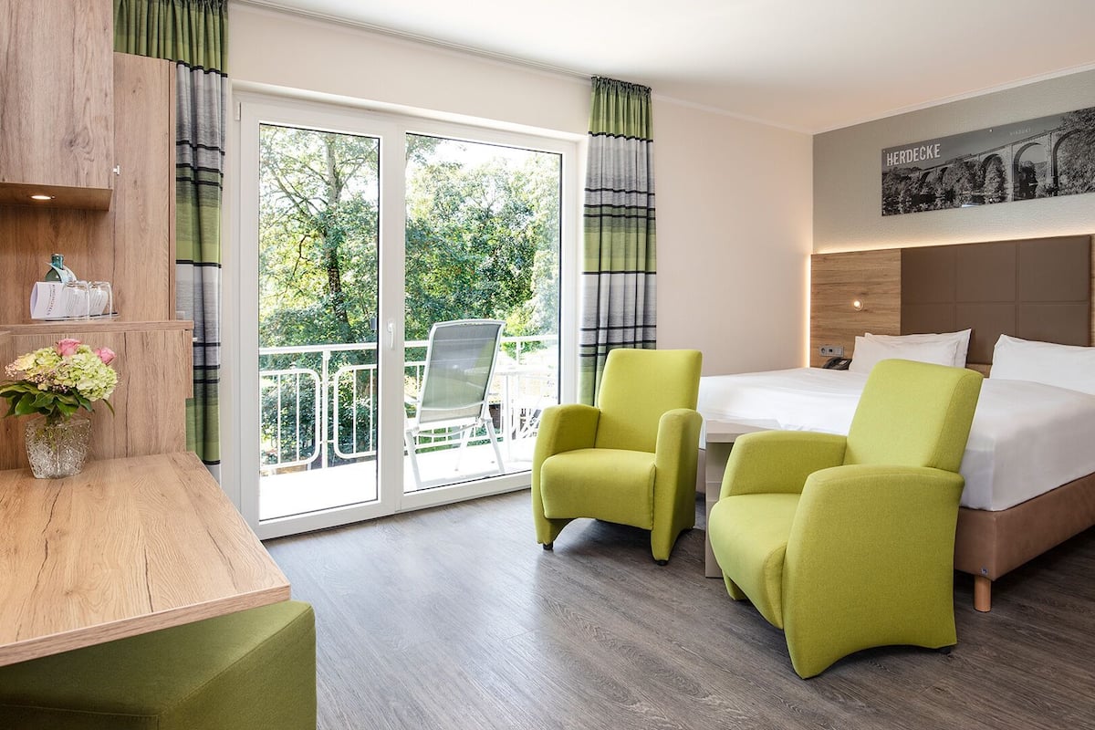 Quarto de hotel com uma cama, duas poltronas verdes, uma escrivaninha de madeira com um vaso de flores e uma varanda com cortinas.