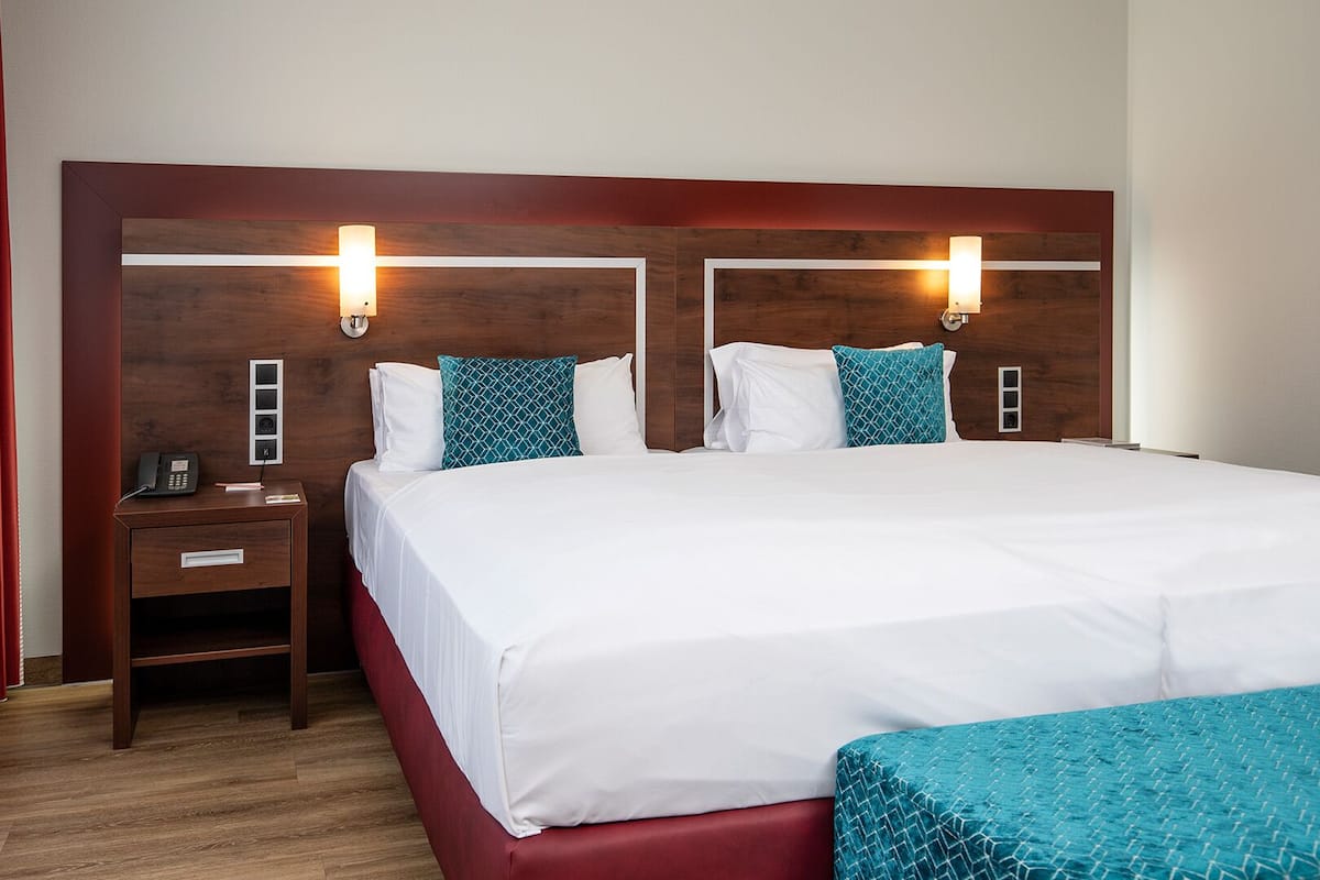 Quarto de hotel com uma cama grande, duas mesas de cabeceira, um criado-mudo e cabeceira de madeira com luminárias fixadas na parede.