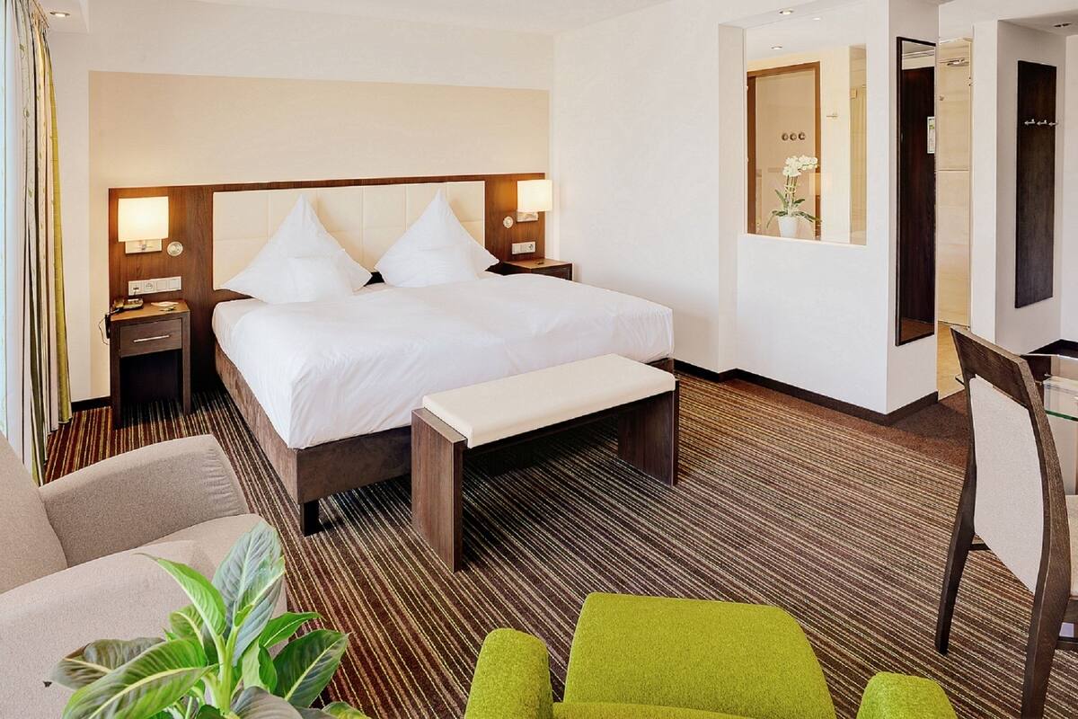 Quarto de hotel com uma cama grande, mesas de cabeceira, um banco, uma cadeira e uma planta em vaso.