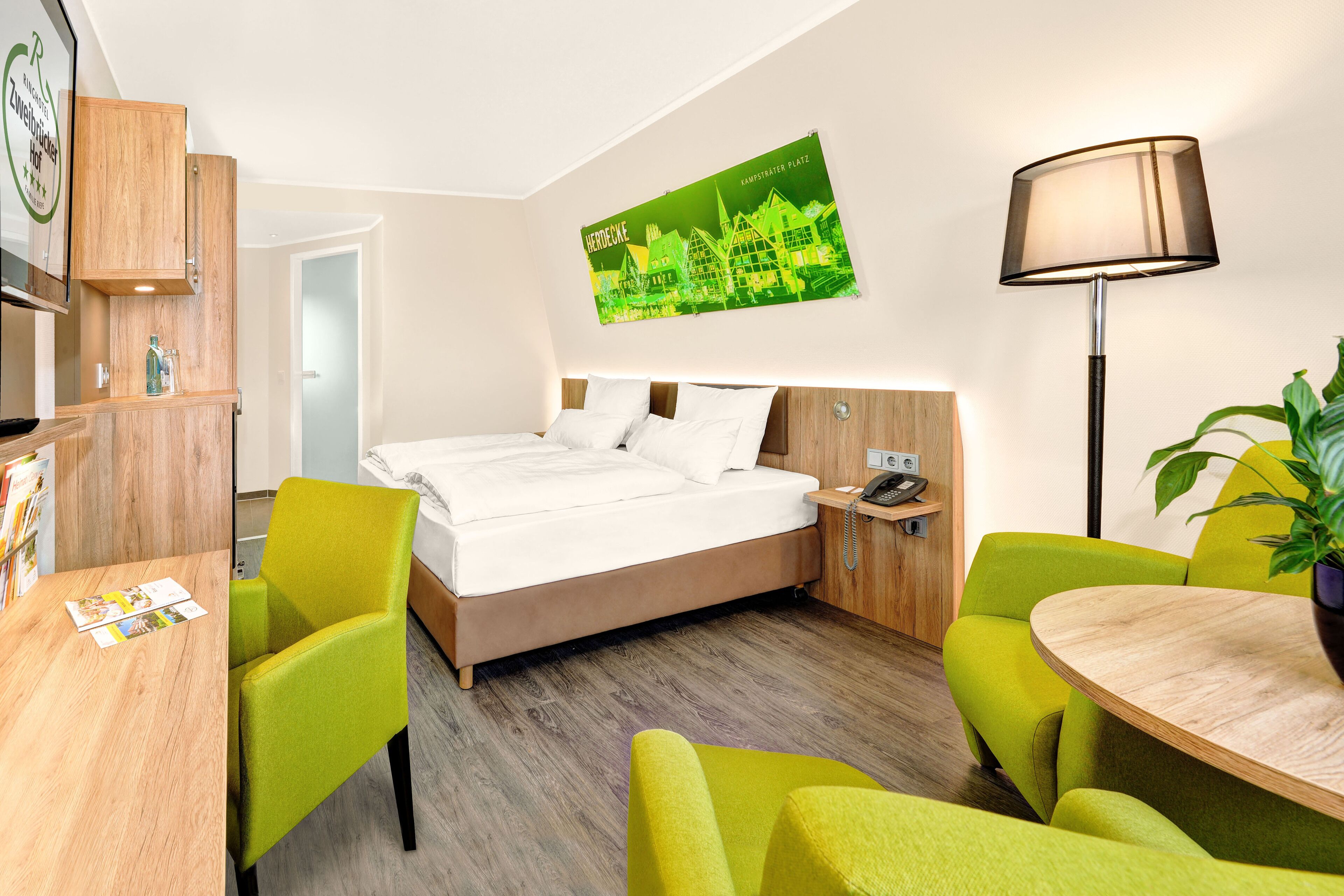 Quarto de hotel com uma cama, uma escrivaninha com cadeira, uma luminária e uma planta.