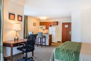 Quarto de hotel compacto com cozinha americana, uma escrivaninha com luminária, uma cadeira e uma cama.