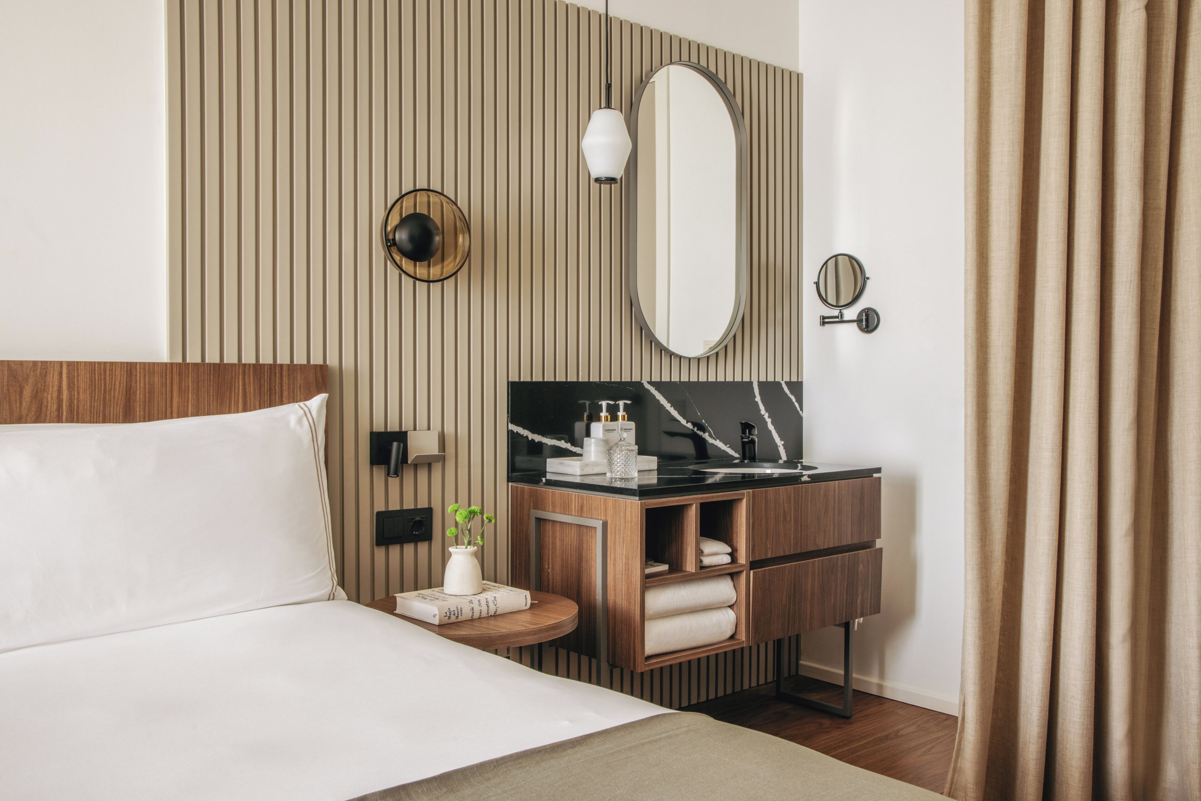 Quarto de hotel com cama, cabeceira de madeira, penteadeira com espelho, mesa de cabeceira redonda e luminária fixa na parede.