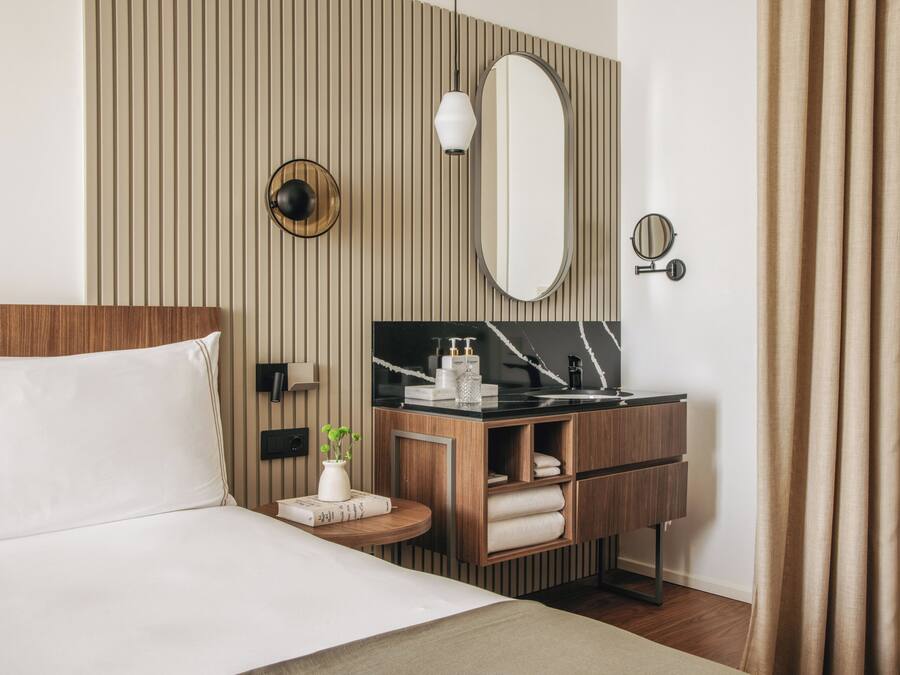 Quarto de hotel com cama, cabeceira de madeira, penteadeira com espelho, mesa de cabeceira redonda e luminária fixa na parede.