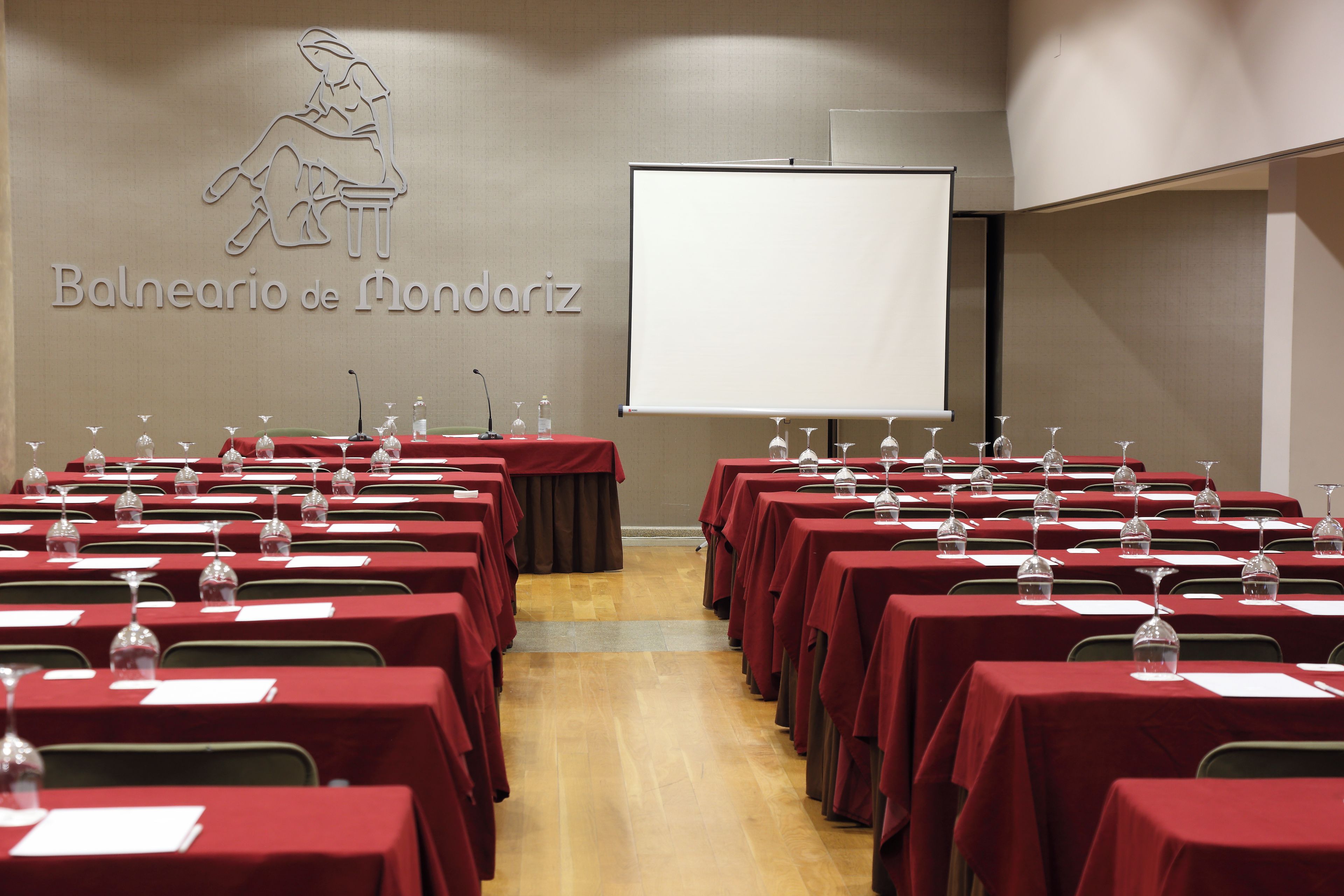 Uma sala de conferências com fileiras de mesas, cada uma com toalha vermelha e um copo sobre elas, e uma tela de projetor na parede.
