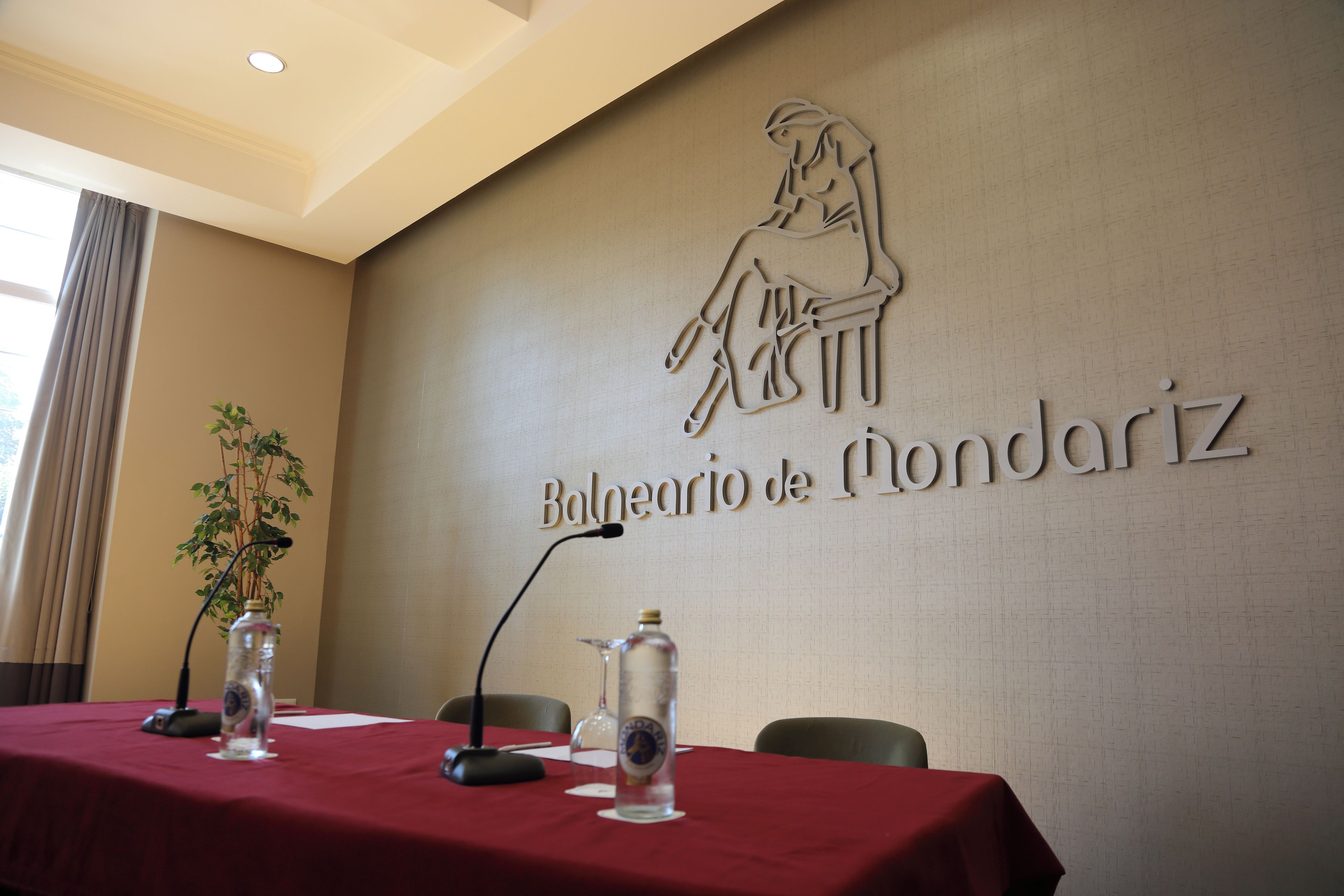 Sala de conferências com uma mesa preparada para uma reunião, um microfone e uma parede decorativa com um logotipo.