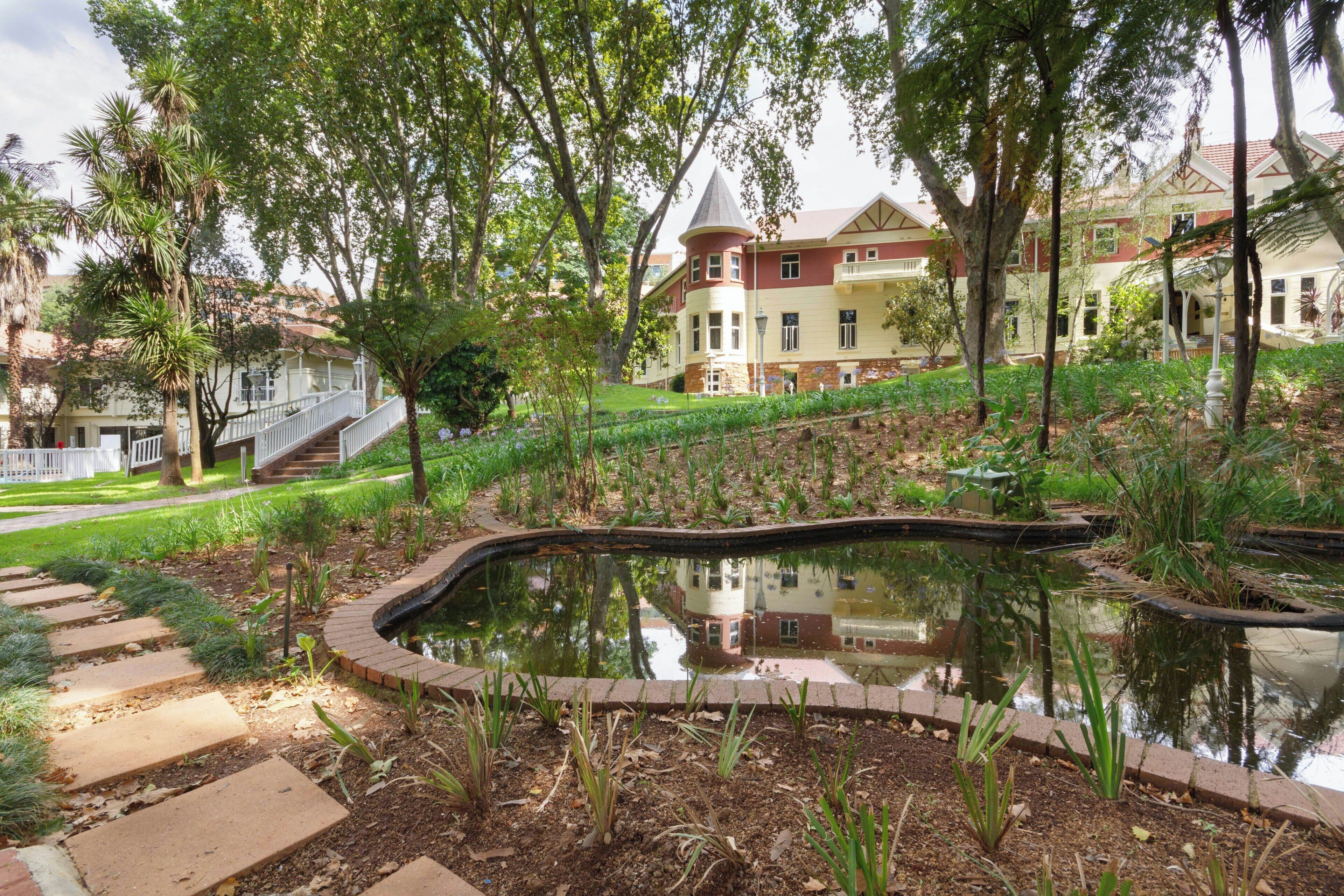 Um jardim paisagístico com lago, caminhos e um edifício com torre ao fundo.