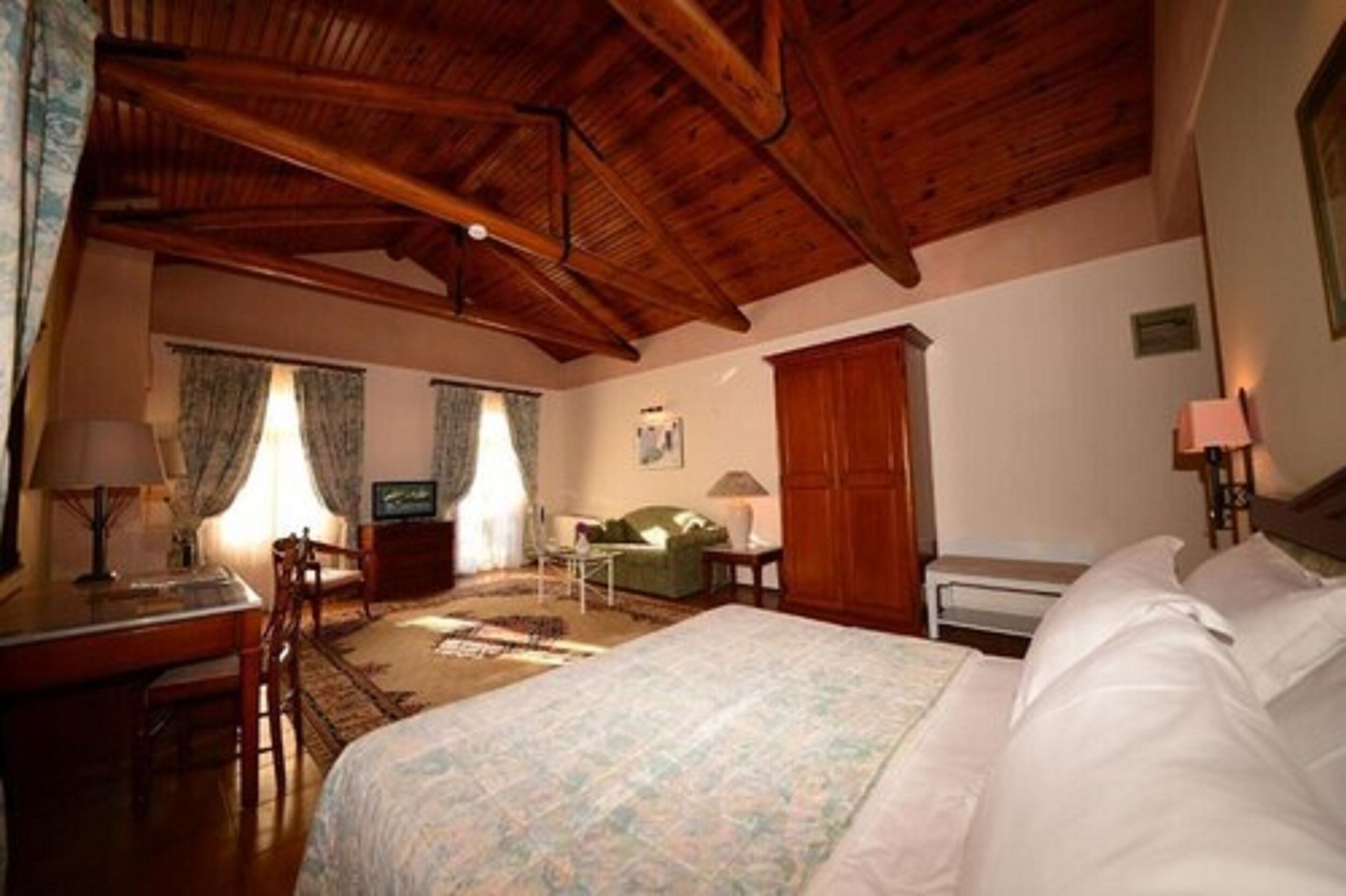 Quarto com teto de madeira, cama com lençóis brancos, escrivaninha com abajur, uma cadeira e uma televisão.