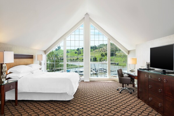 Quarto de hotel com uma cama grande, uma escrivaninha com televisão, uma cadeira e vista para uma marina através de amplas janelas.