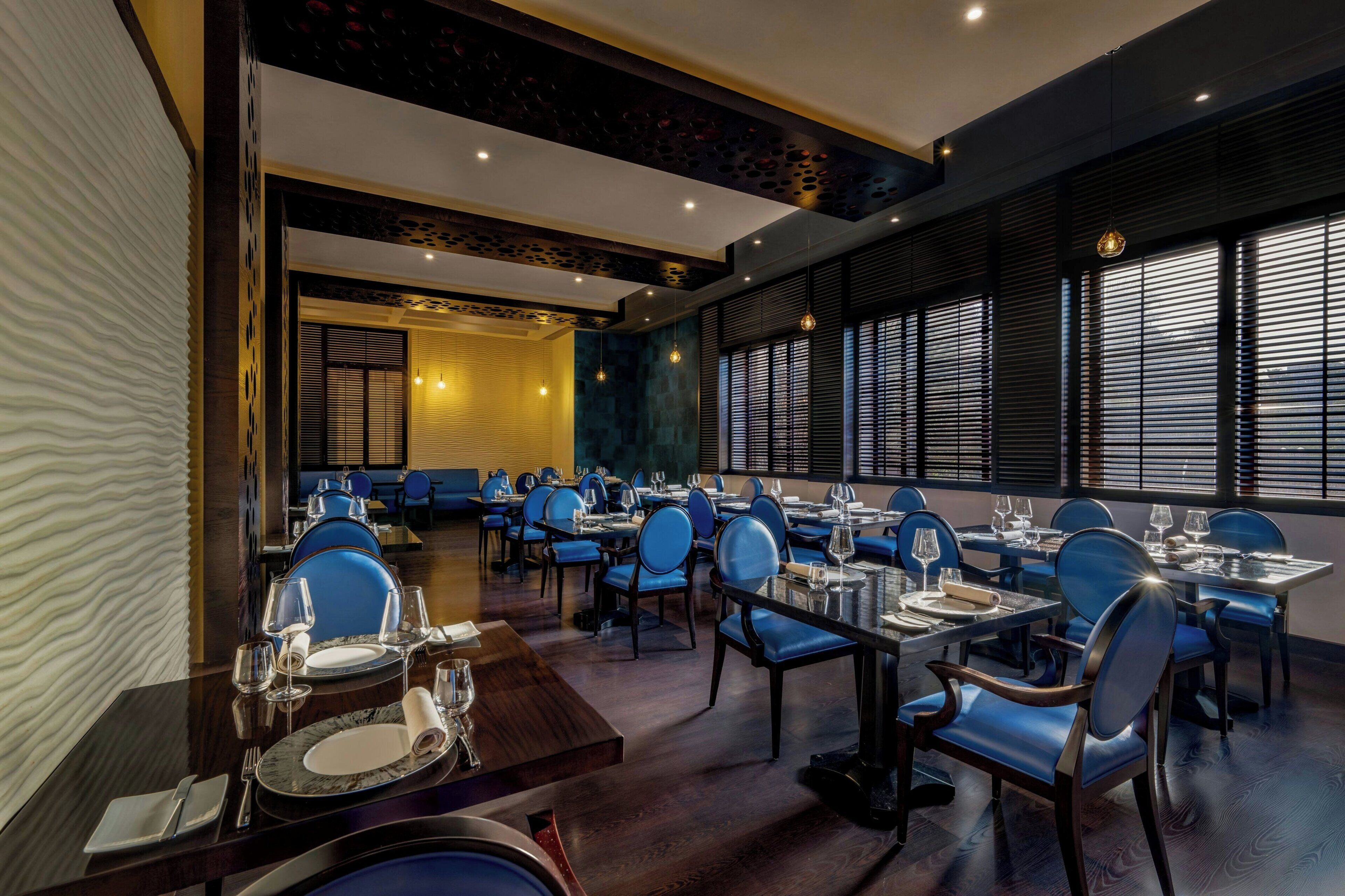 Restaurante com mesas preparadas para refeições, cadeiras azuis e vista para o exterior através de grandes janelas.