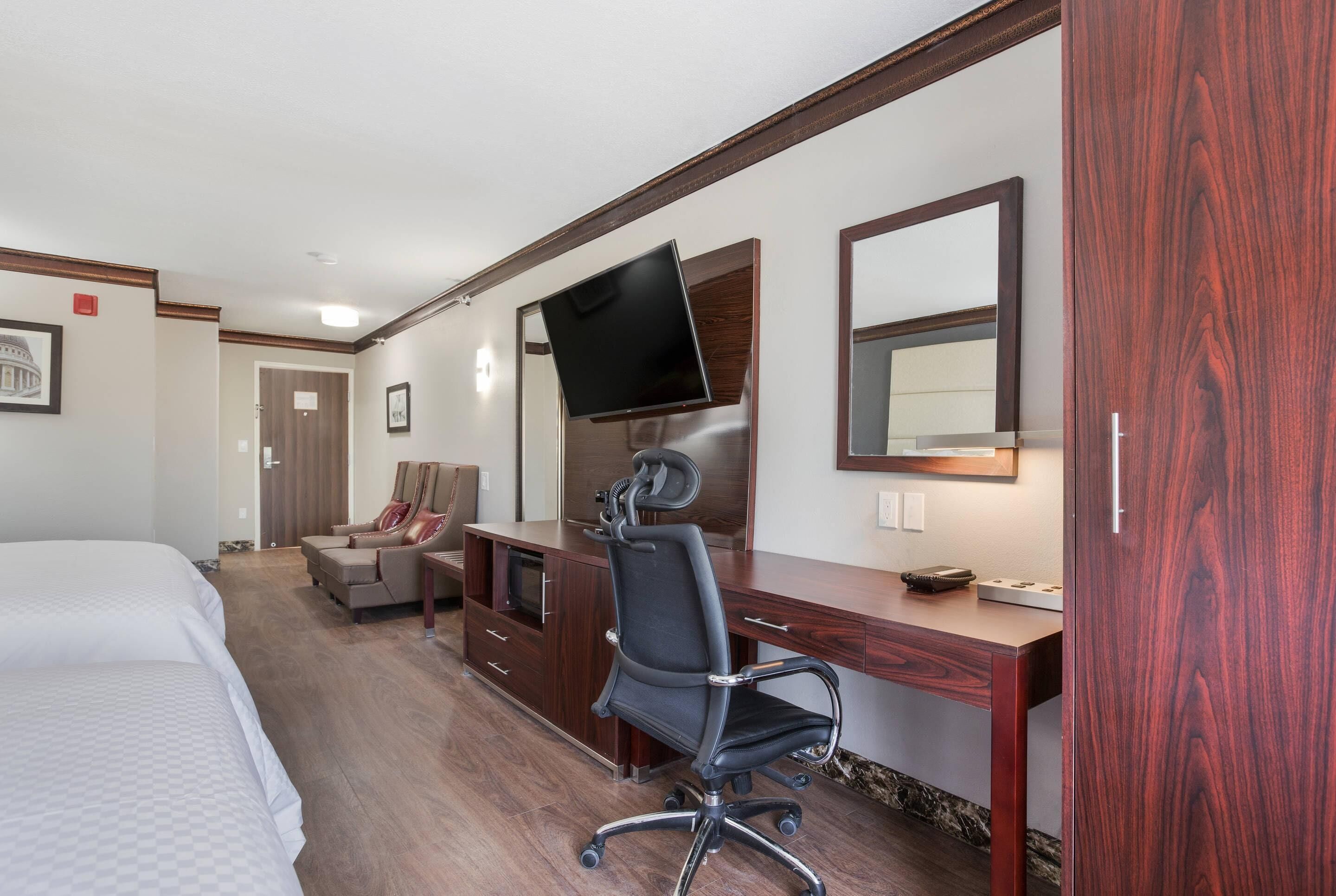 Quarto de hotel com cama, secretária e cadeira, televisão fixada na parede e guarda-roupa de madeira.