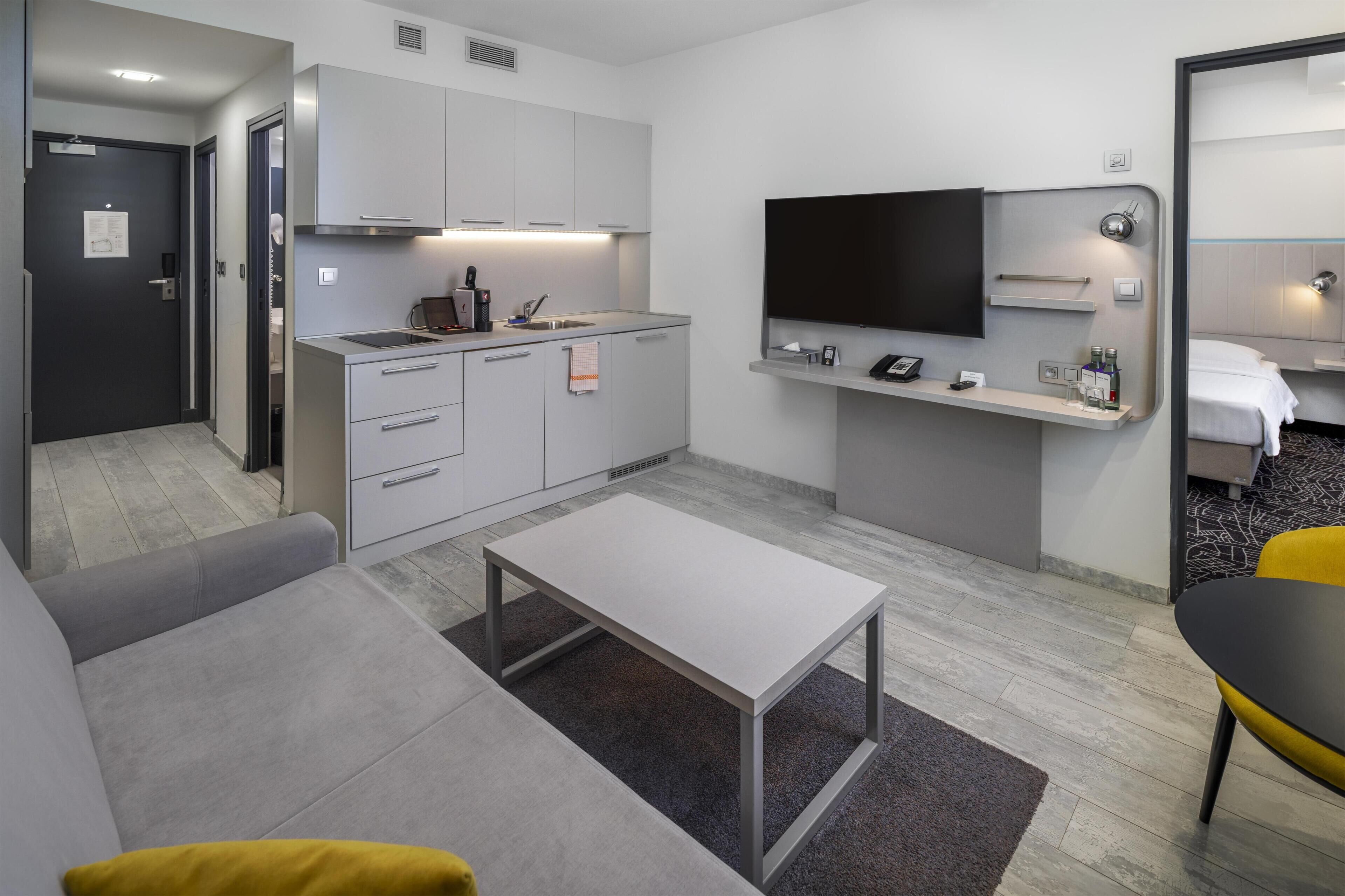 Quarto de hotel moderno com cozinha compacta, televisão de tela plana, sofá, mesa de jantar e uma cama pequena.