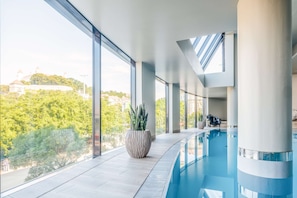 Piscina interior moderna com amplas janelas e vista para áreas verdes e edifícios.