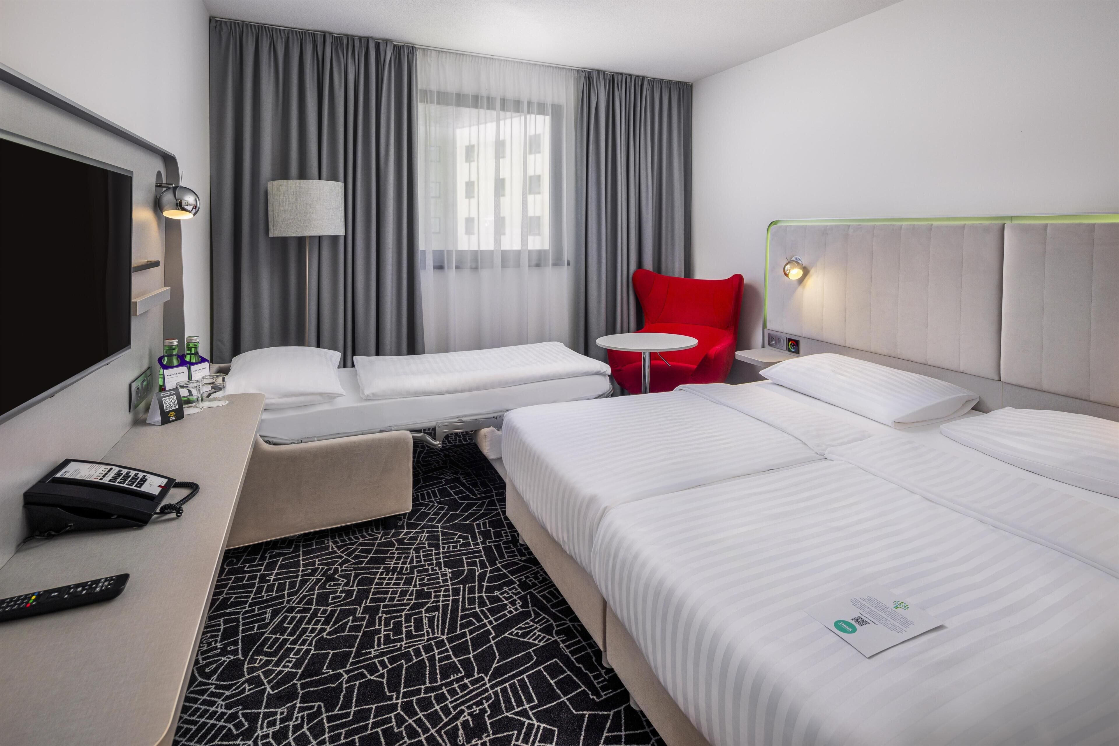 Quarto de hotel com cama, escrivaninha, cadeira, televisão e janela com cortinas.