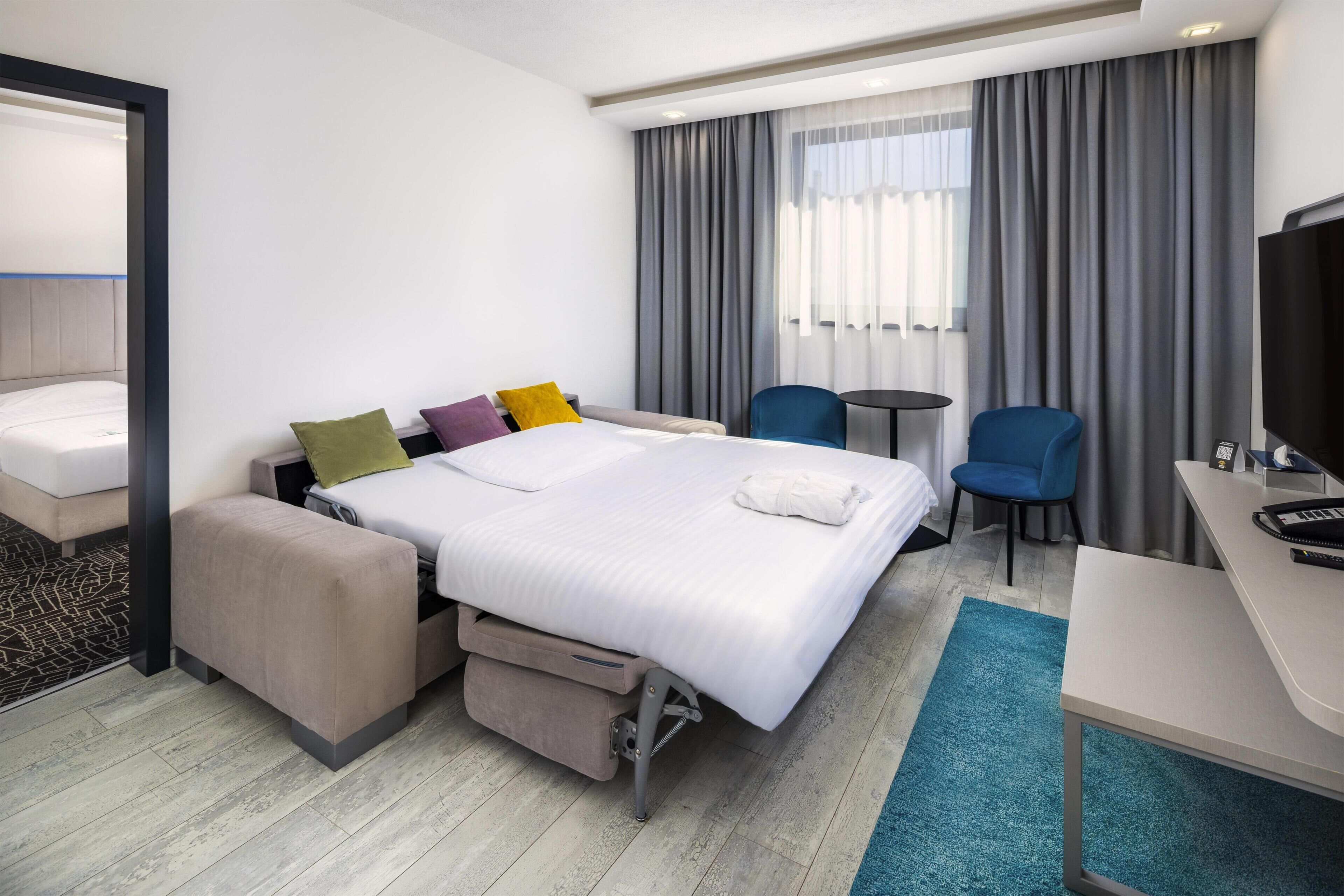 Quarto de hotel com cama, escrivaninha, cadeira, televisão e janela com cortinas.