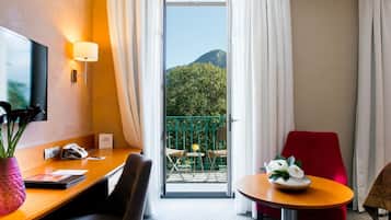 Quarto de hotel com varanda, escrivaninha, cadeira, televisão e mesa com um vaso de flores.