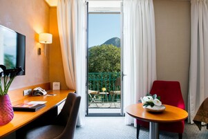 Quarto de hotel com varanda, escrivaninha, cadeira, televisão e mesa com um vaso de flores.
