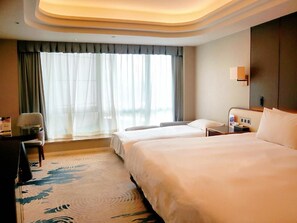 Quarto de hotel com duas camas, uma escrivaninha, uma cadeira e uma janela grande com cortinas.